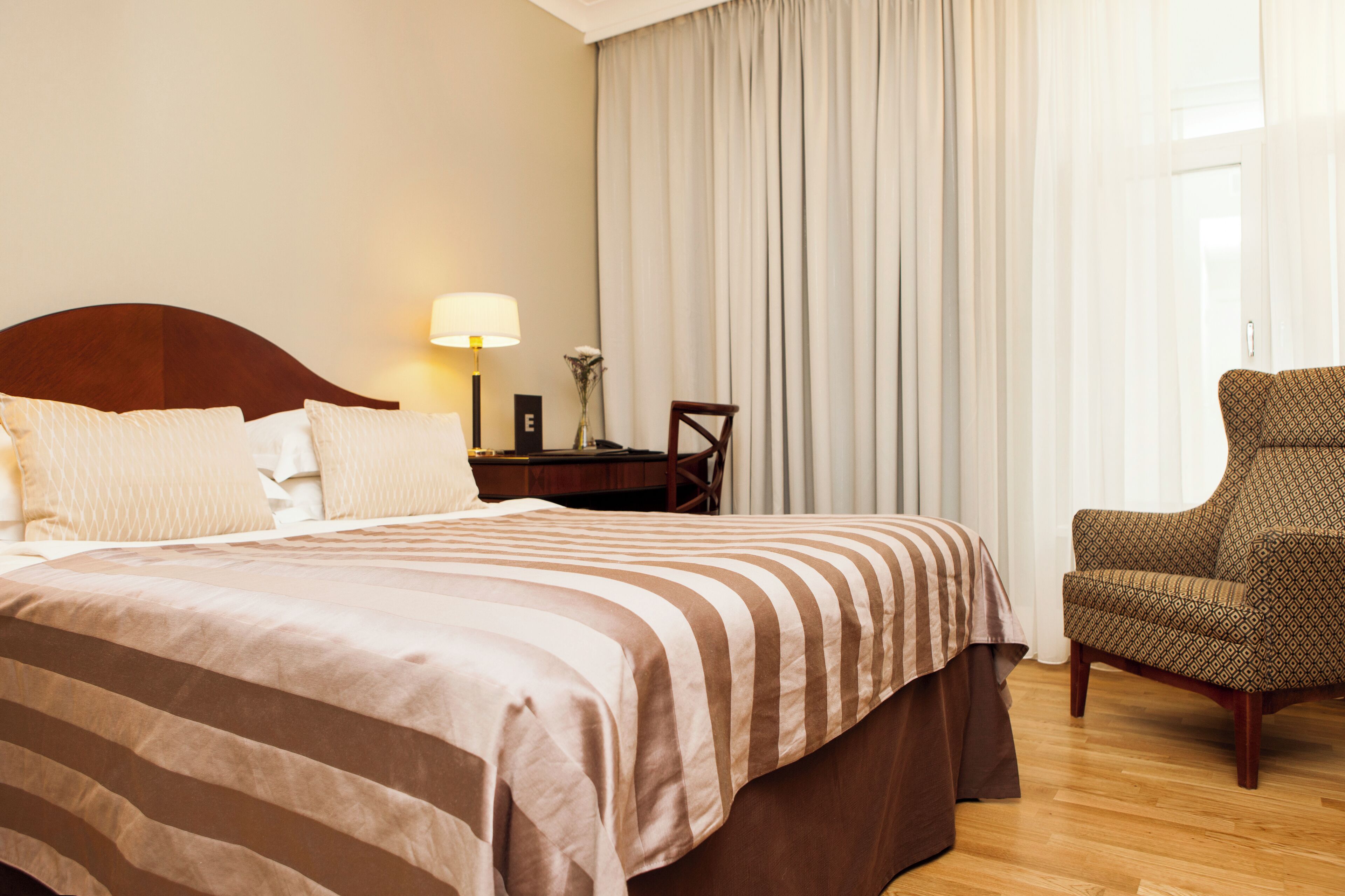 Quarto de hotel com uma cama grande, uma mesa com luminária, uma cadeira e uma janela com cortinas.