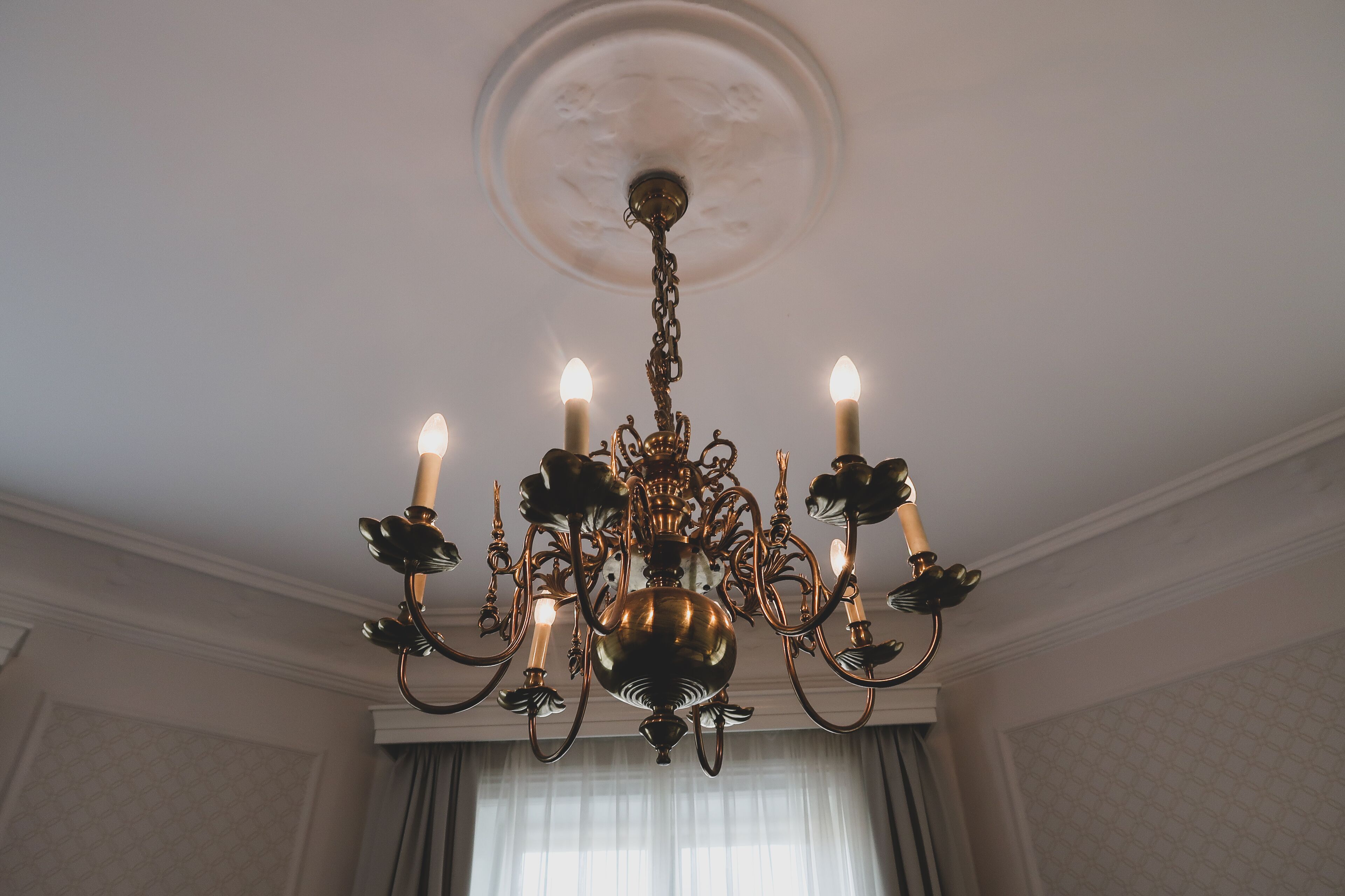 Um candelabro com várias luzes semelhantes a velas, um teto decorativo e uma janela com cortinas.