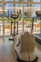 Uma academia com equipamentos de ginástica, incluindo uma elíptica e uma esteira, e vista para área de estar externa e palmeiras através de grandes janelas.