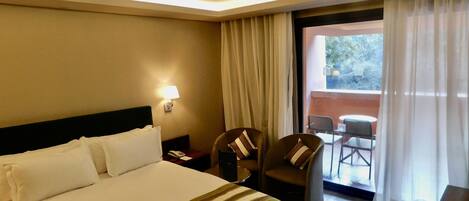 Quarto de hotel com uma cama grande, mesa de cabeceira com telefone, uma cadeira e vista para área externa com mesa e cadeiras.