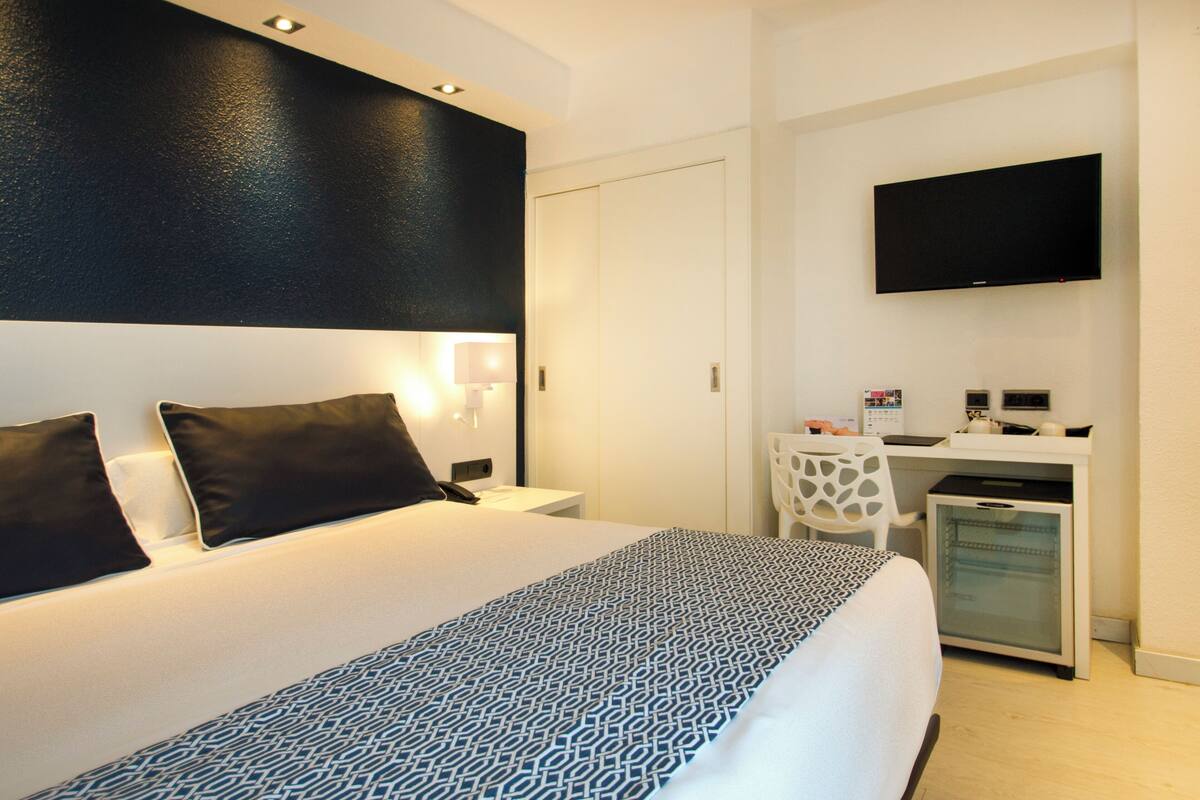Quarto de hotel com cama grande, escrivaninha com computador, televisão e minibar.