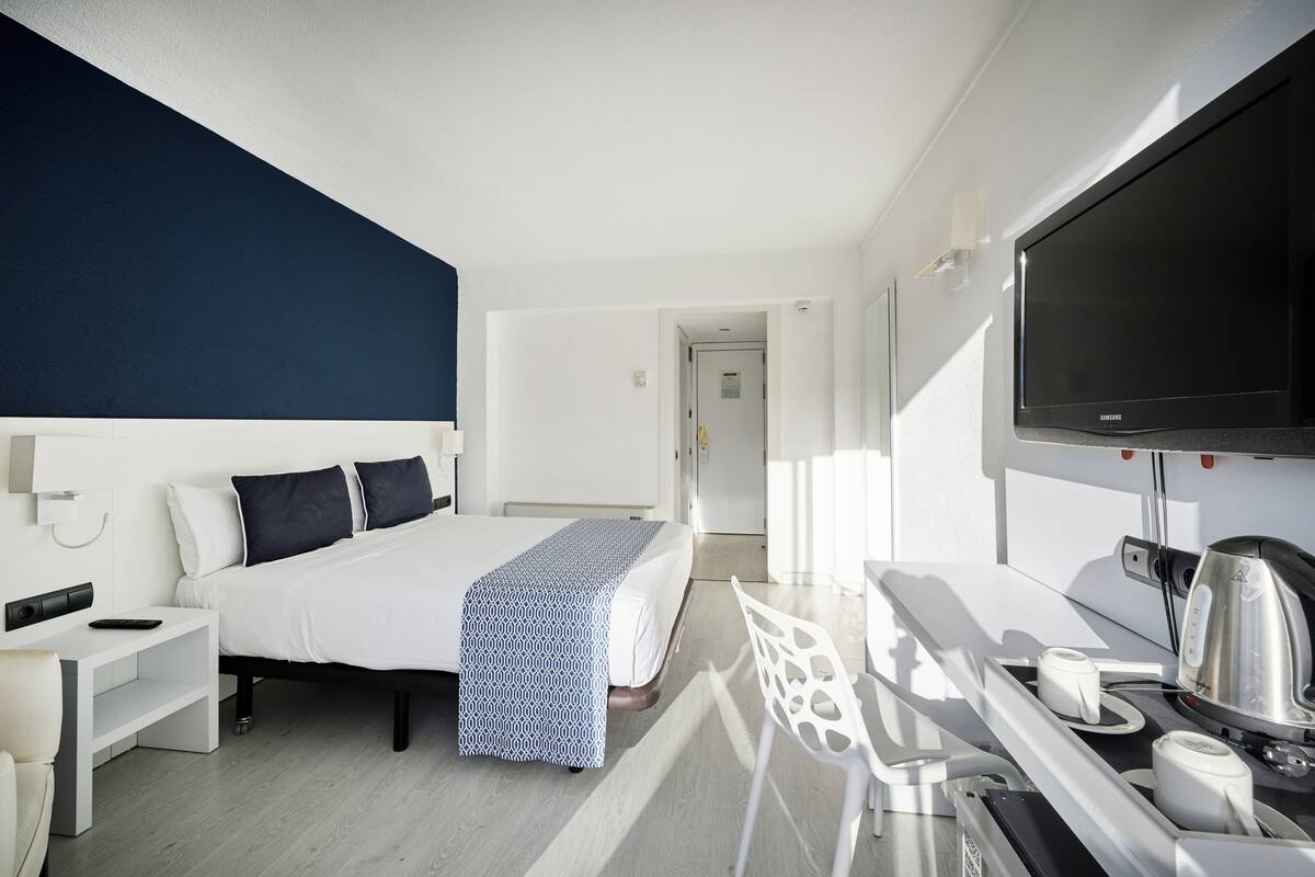 Quarto de hotel moderno com cama, mesa de cabeceira, televisão e área de kitchenette.