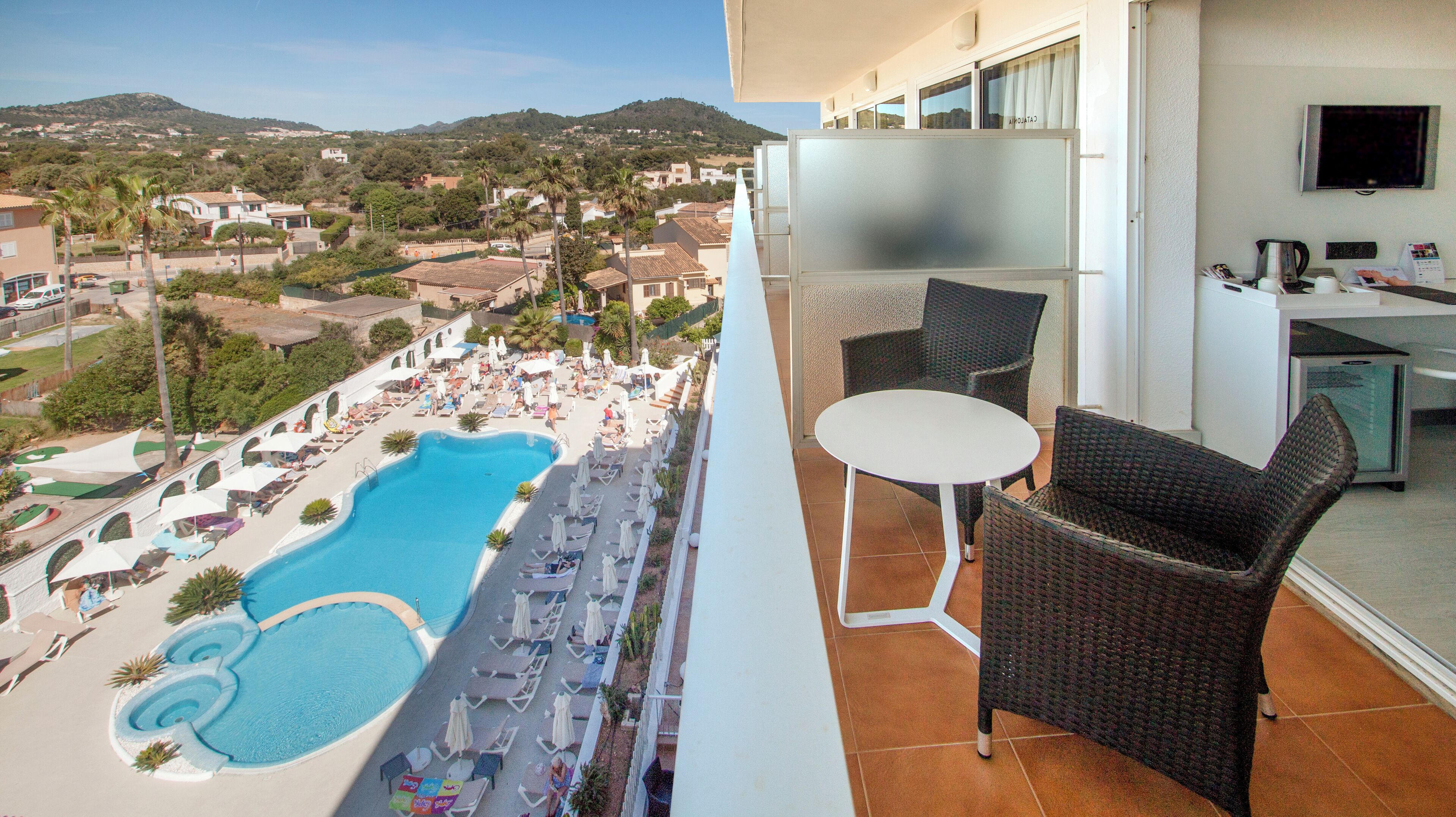 Uma varanda com vista para uma piscina e área de resort, mobiliário de vime e uma televisão de tela plana.