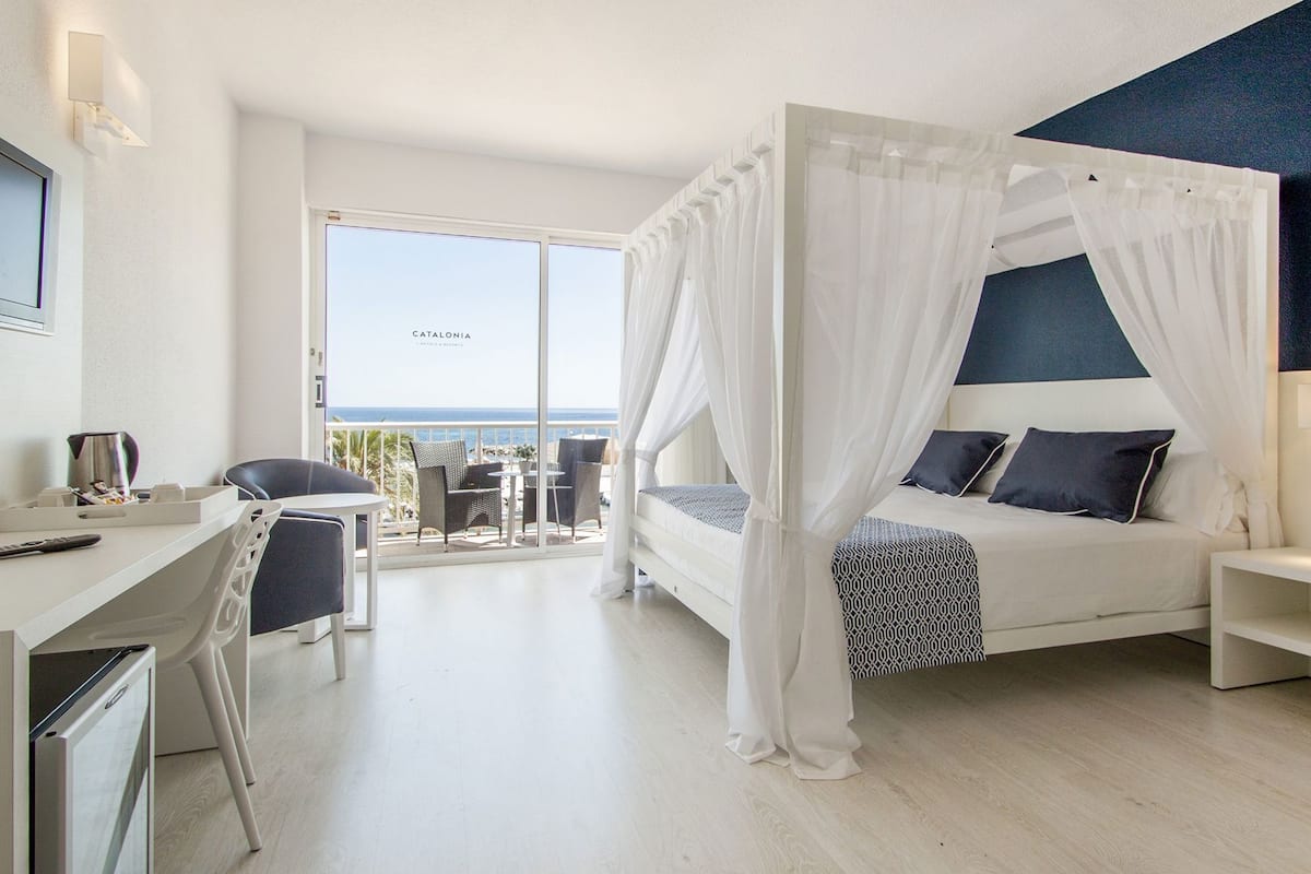 Quarto com cama de dossel, uma escrivaninha com cadeira, uma pequena mesa com cadeiras e vista para o mar.