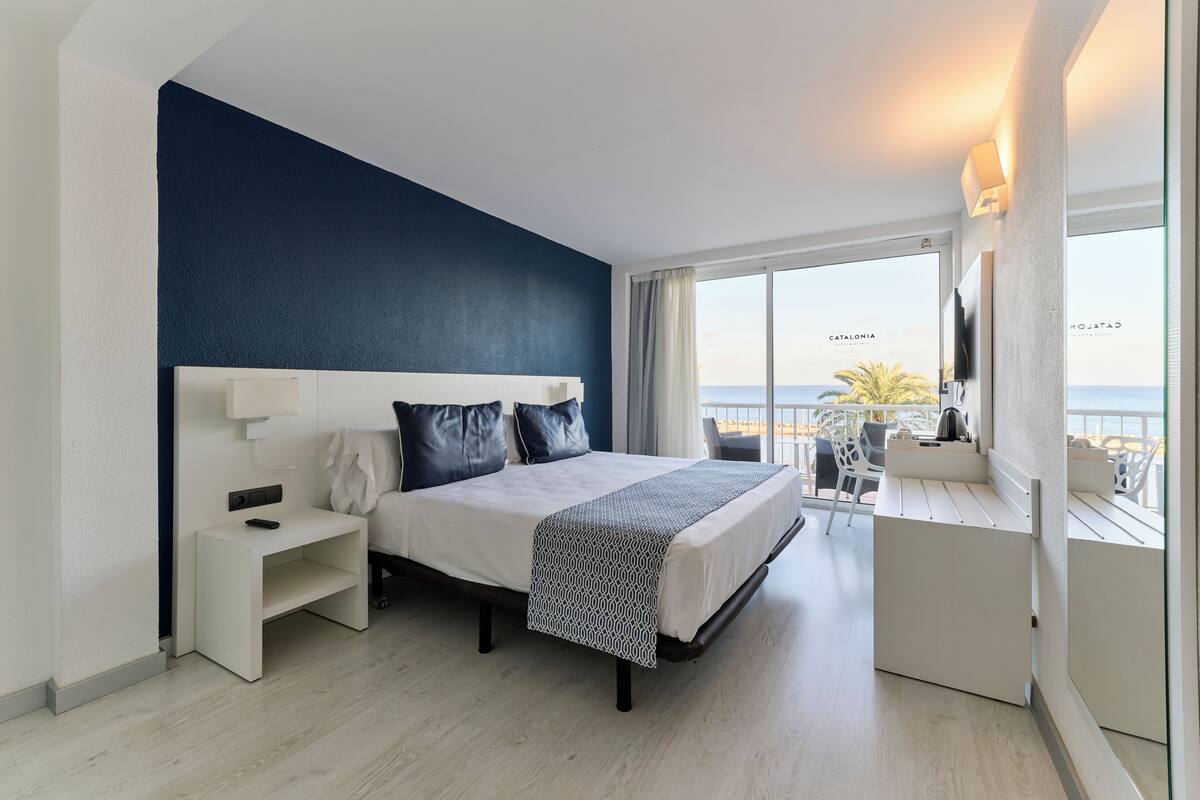 Quarto de hotel moderno com uma cama grande, uma mesa de cabeceira, uma TV e uma varanda com vista para o mar.