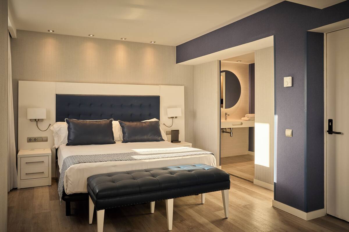 Quarto moderno com uma cama grande, mesas de cabeceira, um banco e um banheiro visível por uma porta aberta.