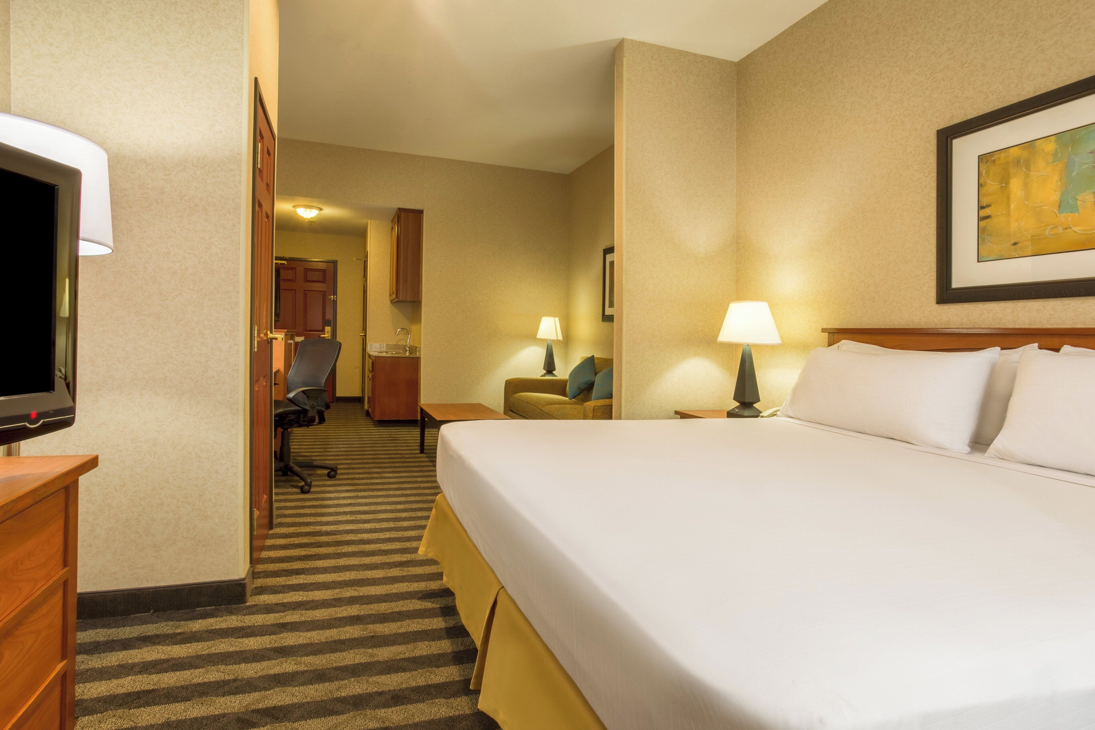 Quarto de hotel com cama, escrivaninha, cadeira, televisão e um quadro na parede.