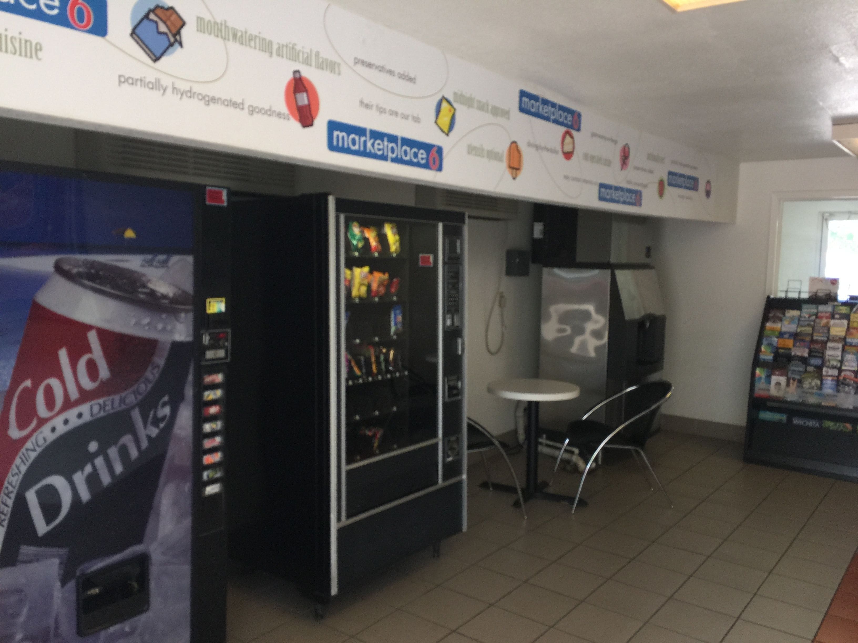 Uma área de máquinas de venda automática com um aviso 'Bebidas Geladas', um aviso 'Mercado' e uma pequena mesa com cadeiras.