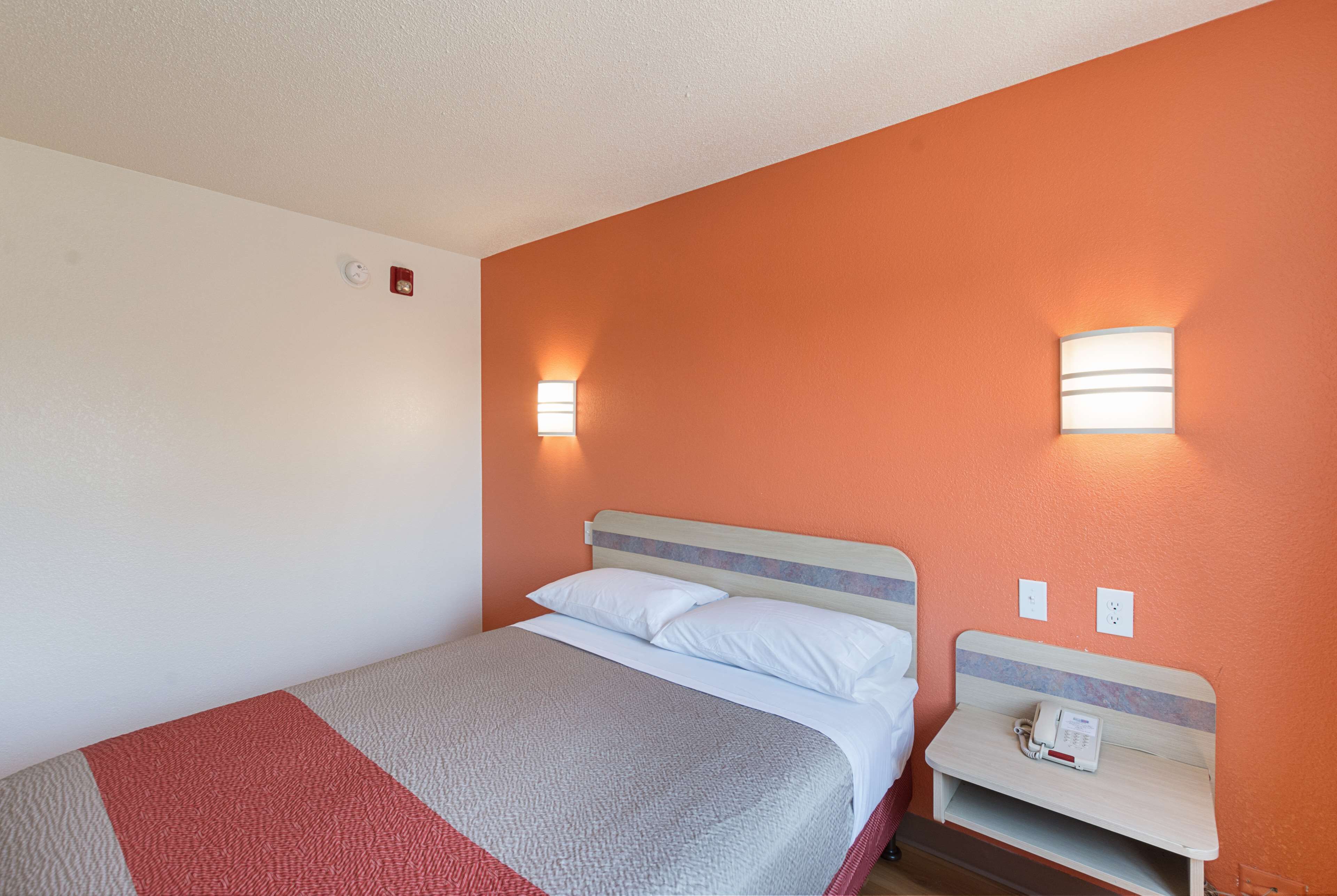 Quarto de hotel com uma cama, mesa de cabeceira com telefone e duas luminárias na parede.