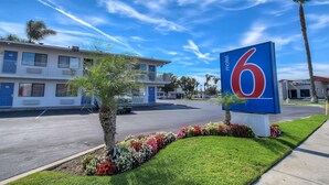 Um motel com um grande letreiro azul indicando a sala número 6, estacionamento e um gramado bem cuidado com flores coloridas.