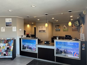 Bancada de recepção de hotel com tela digital exibindo uma mensagem de reserva, suporte para revistas de viagem e uma obra de arte com paisagem urbana emoldurada.