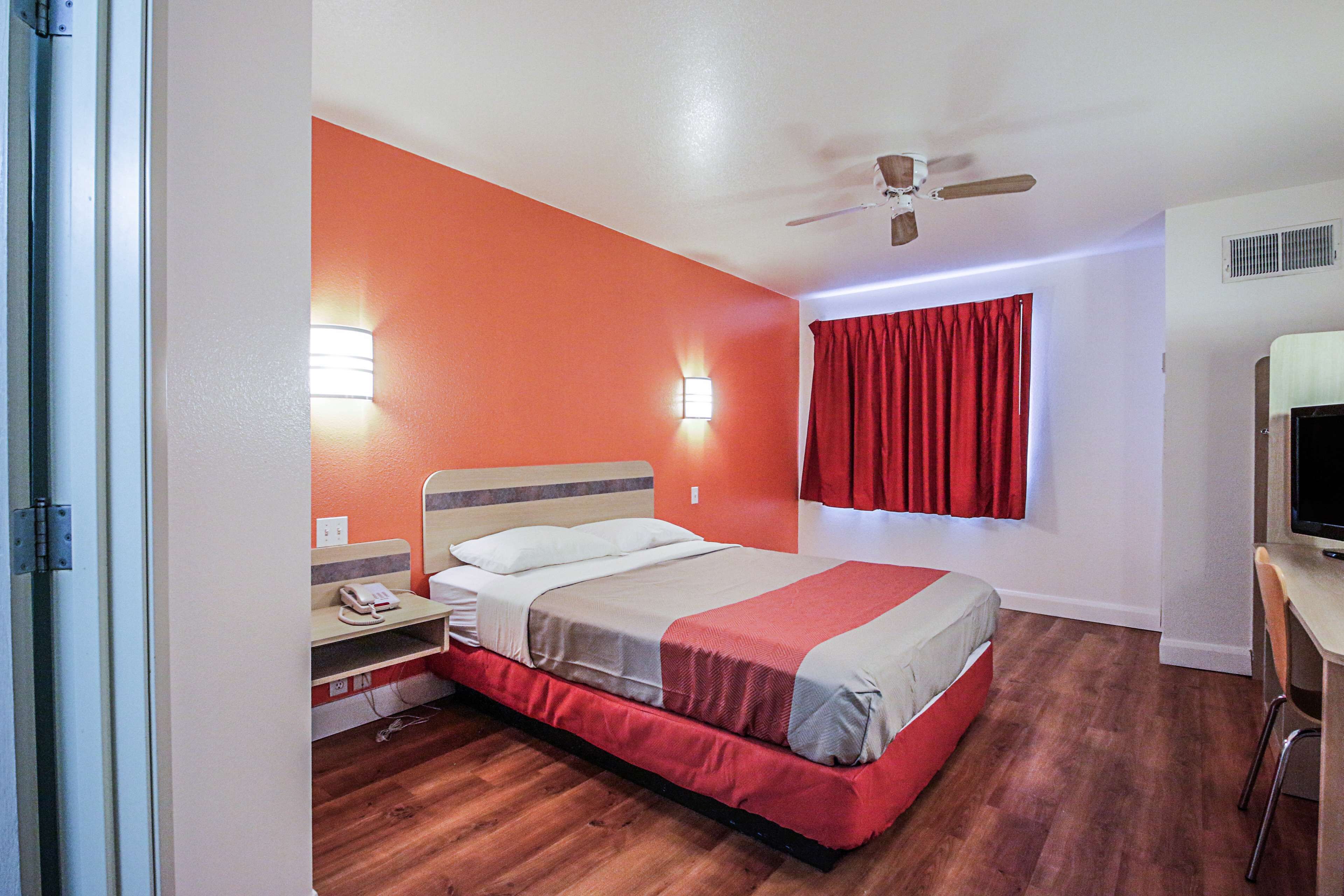 Quarto de hotel com cama, escrivaninha com cadeira, ventilador de teto e janela com cortinas vermelhas.