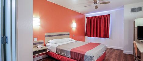 Quarto de hotel com cama, escrivaninha com cadeira, ventilador de teto e janela com cortinas vermelhas.