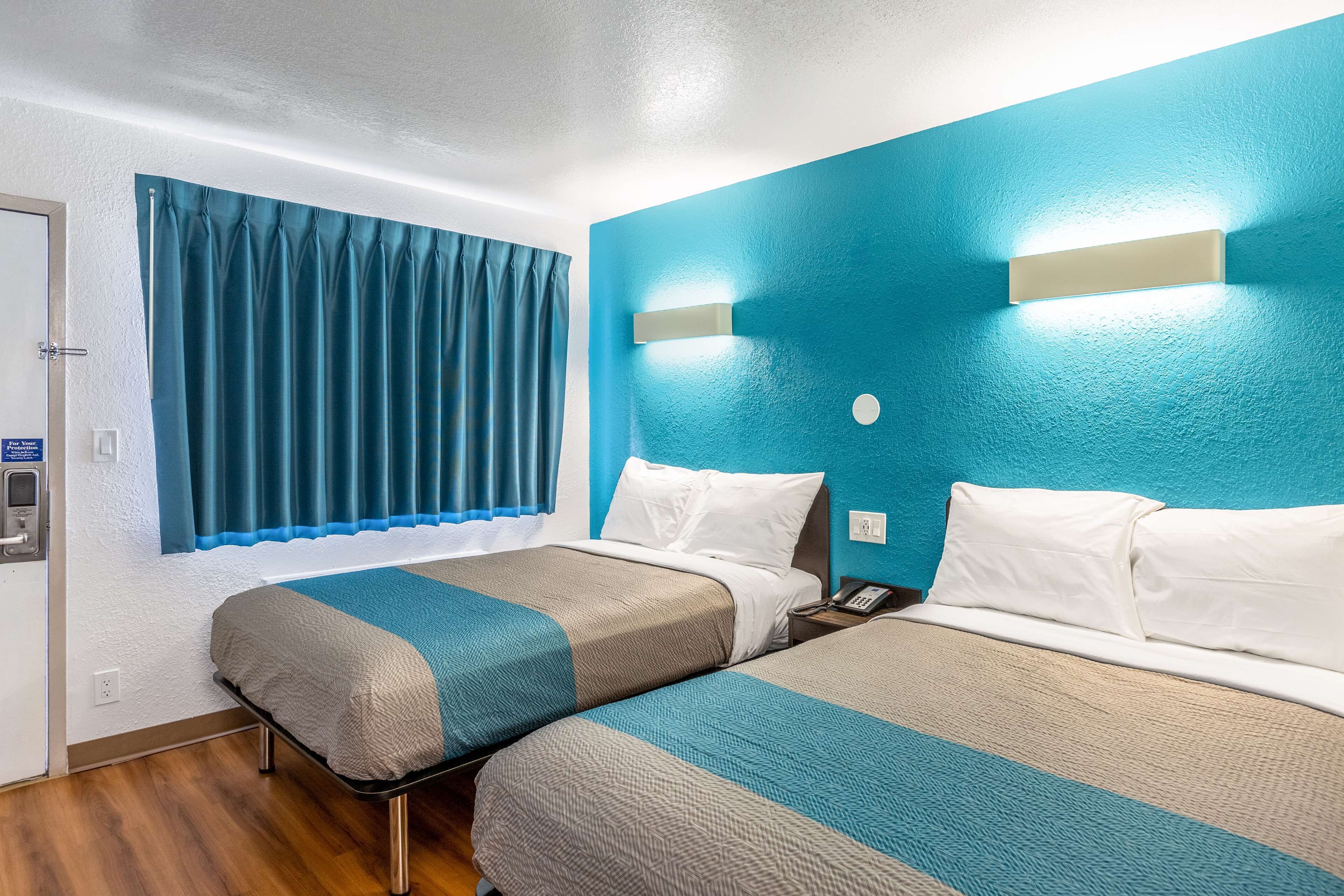 Quarto de hotel com duas camas, uma janela com cortinas azuis, telefone fixado na parede e interruptor de luz.