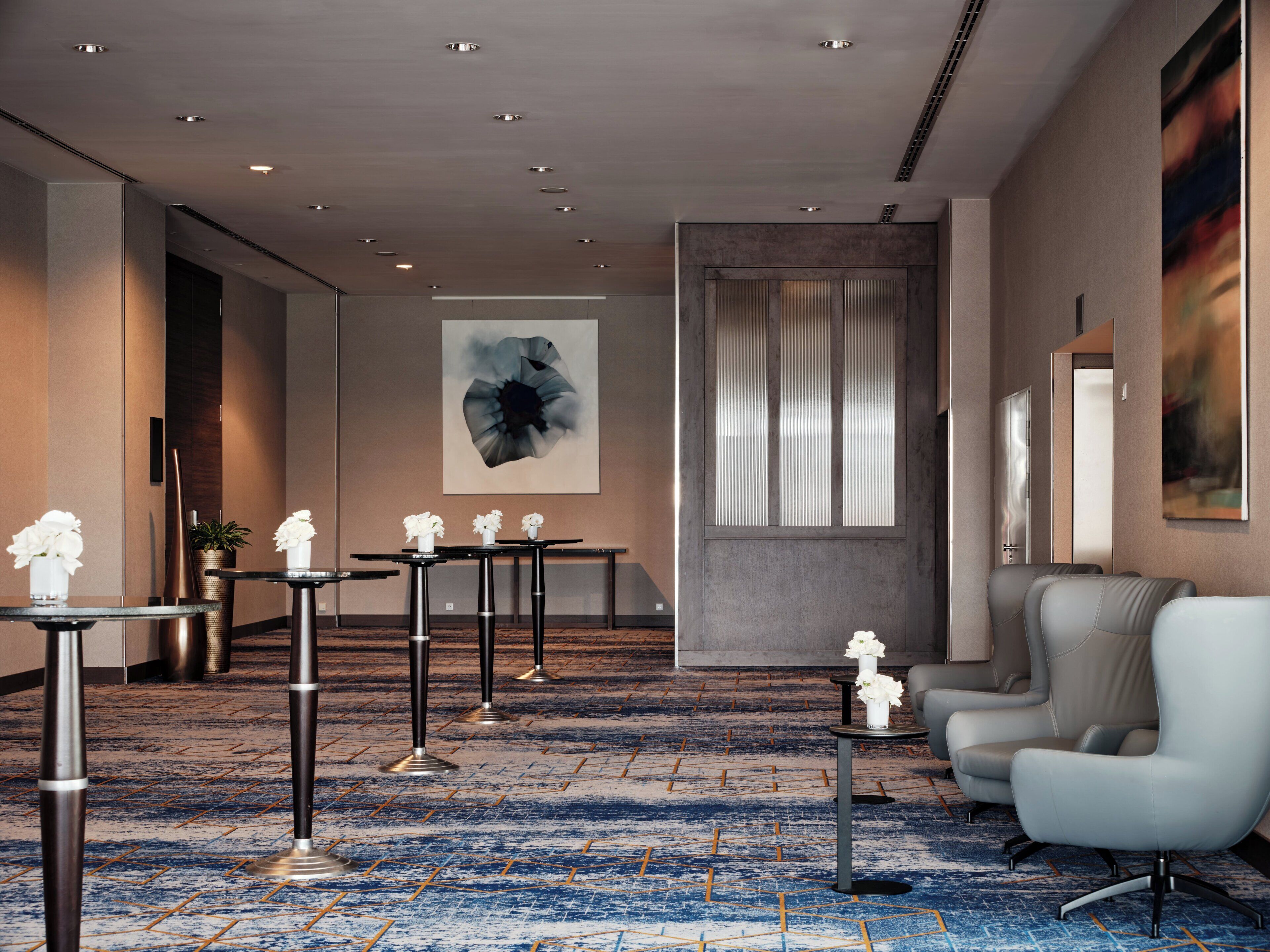 Um saguão de hotel com carpete padronado em azul, área de estar com uma poltrona cinza, uma mesa com um vaso de flores e uma grande pintura abstrata na parede.