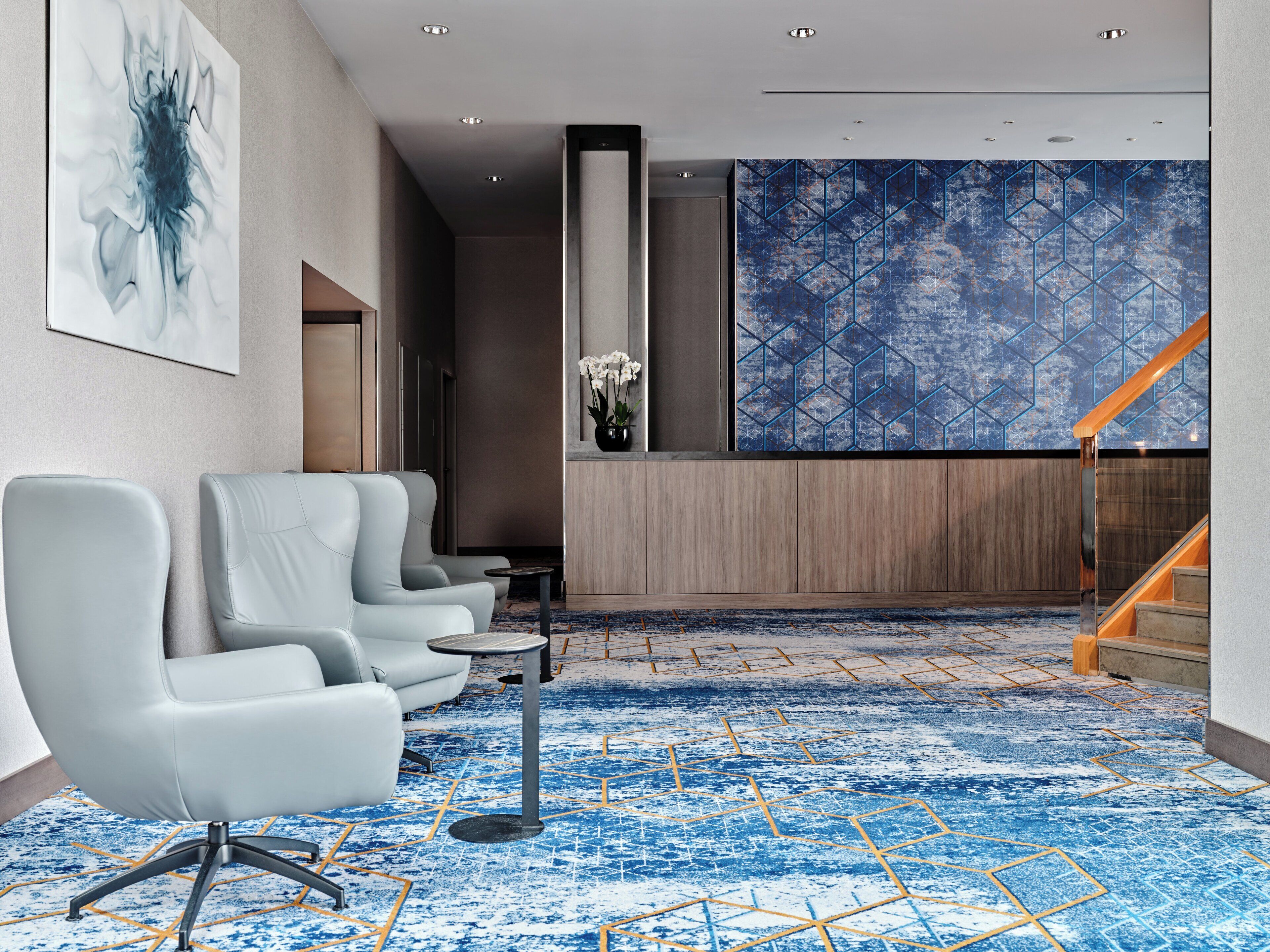 Um lobby moderno com carpete padronado em azul, dois poltronas cinzas, um balcão de recepção e uma escadaria.