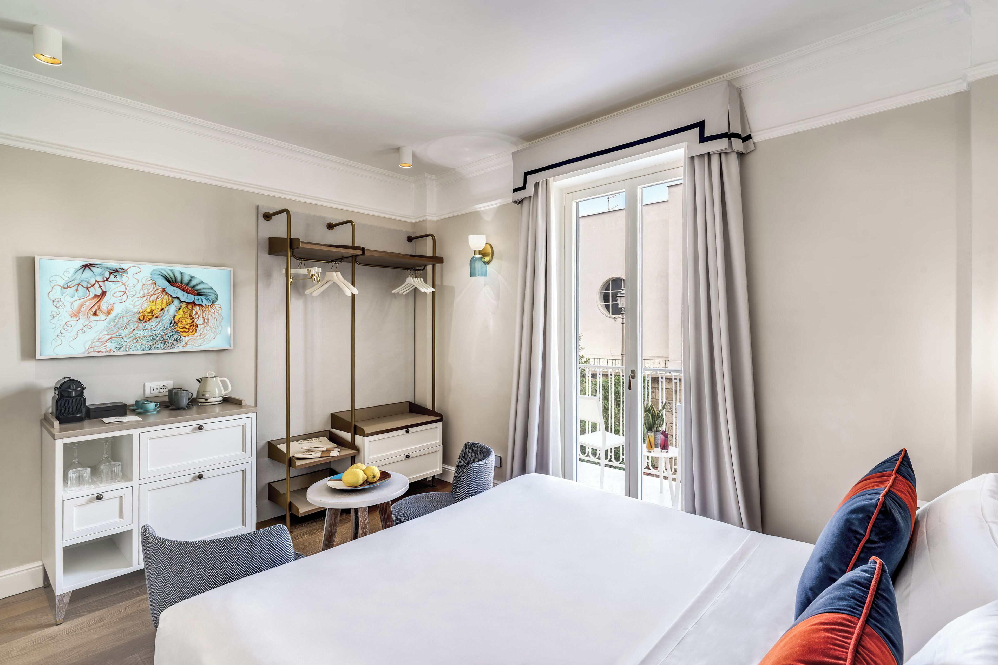 Quarto de hotel com cama, escrivaninha, cadeira, mesinha com frutas e varanda com cortinas.