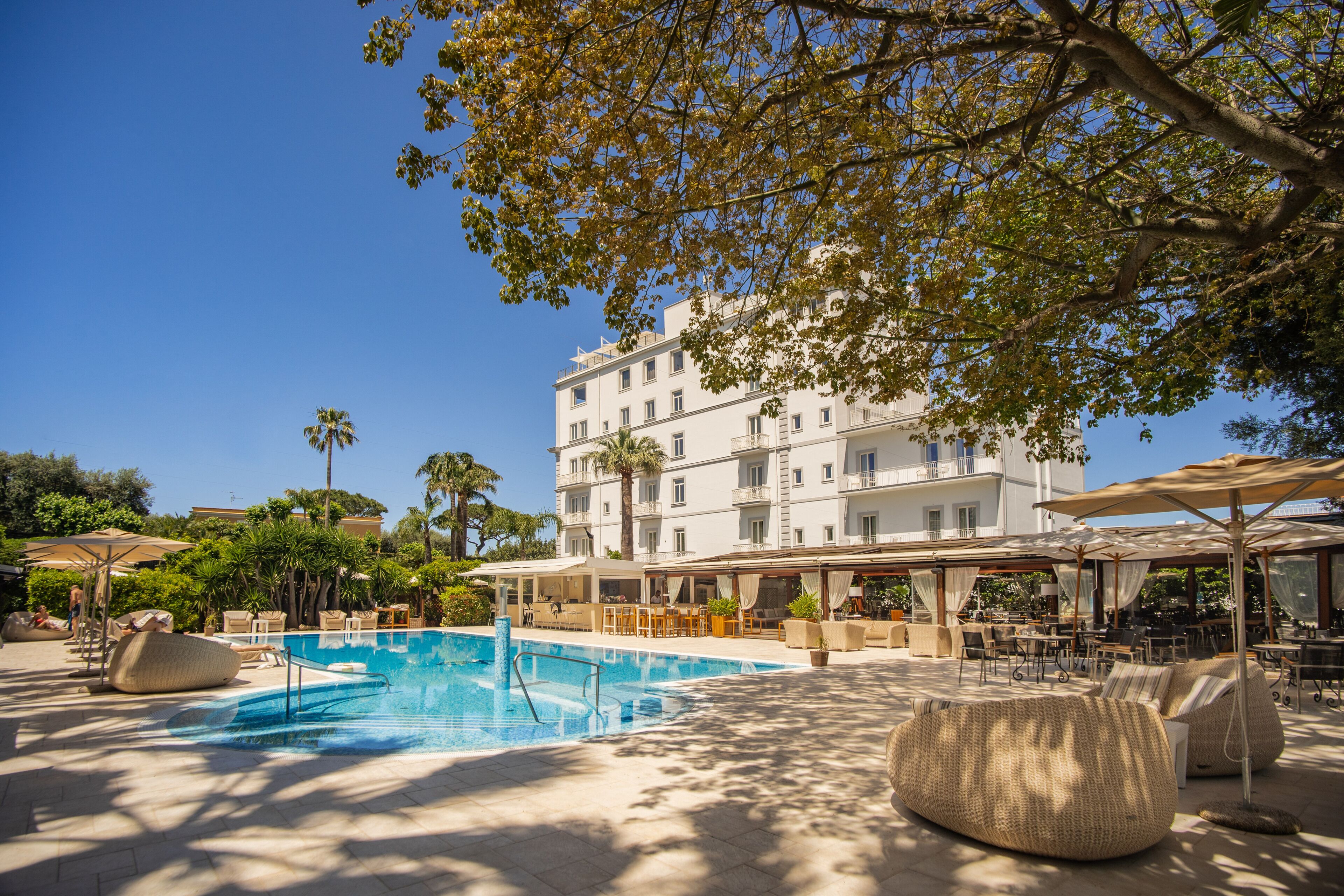 Uma área de piscina com espreguiçadeiras e guarda-sóis, rodeada por um edifício com vários andares e varandas.