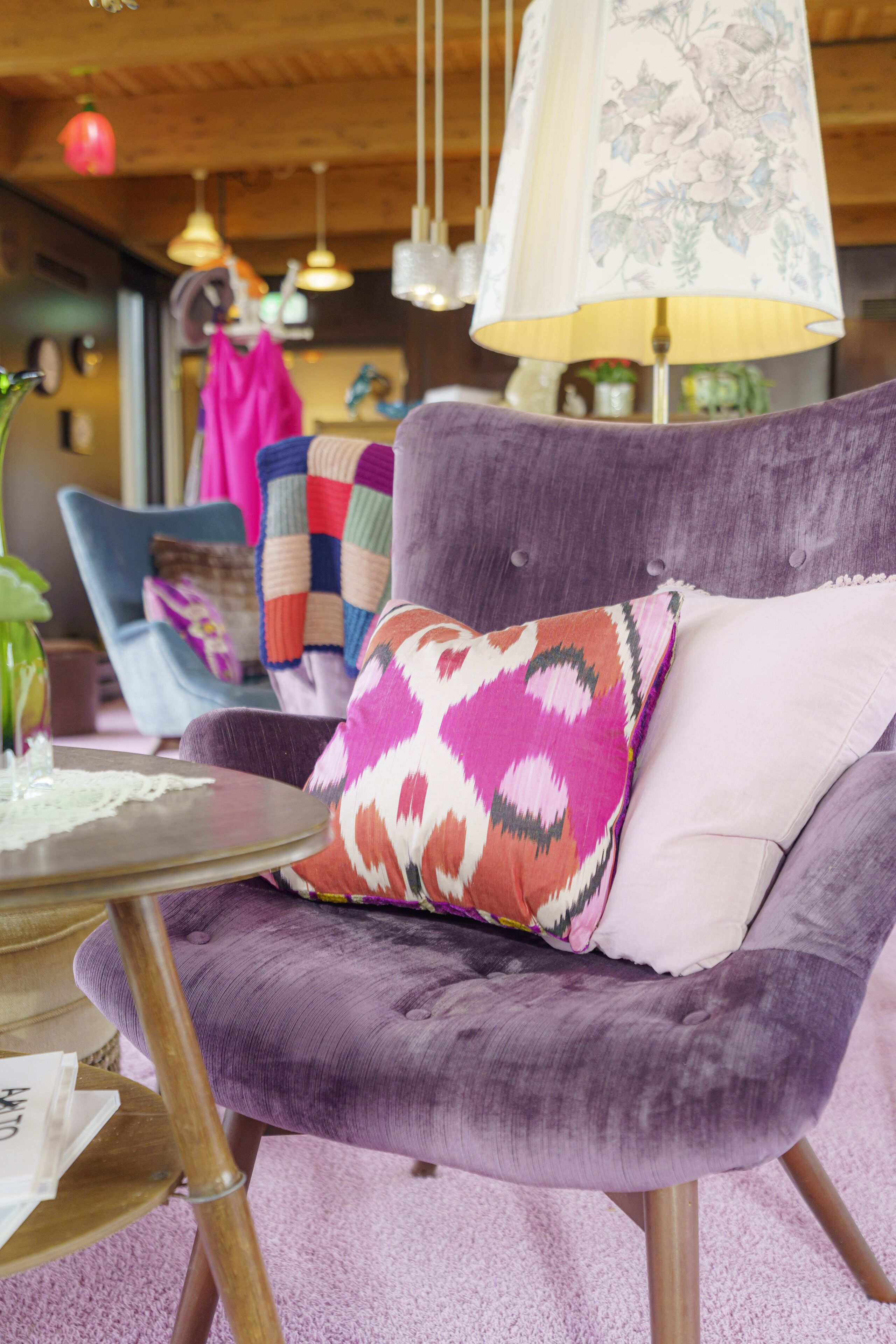 Uma poltrona roxa com almofada estampada, uma mesa de madeira e um candeeiro pendente com motivos florais.