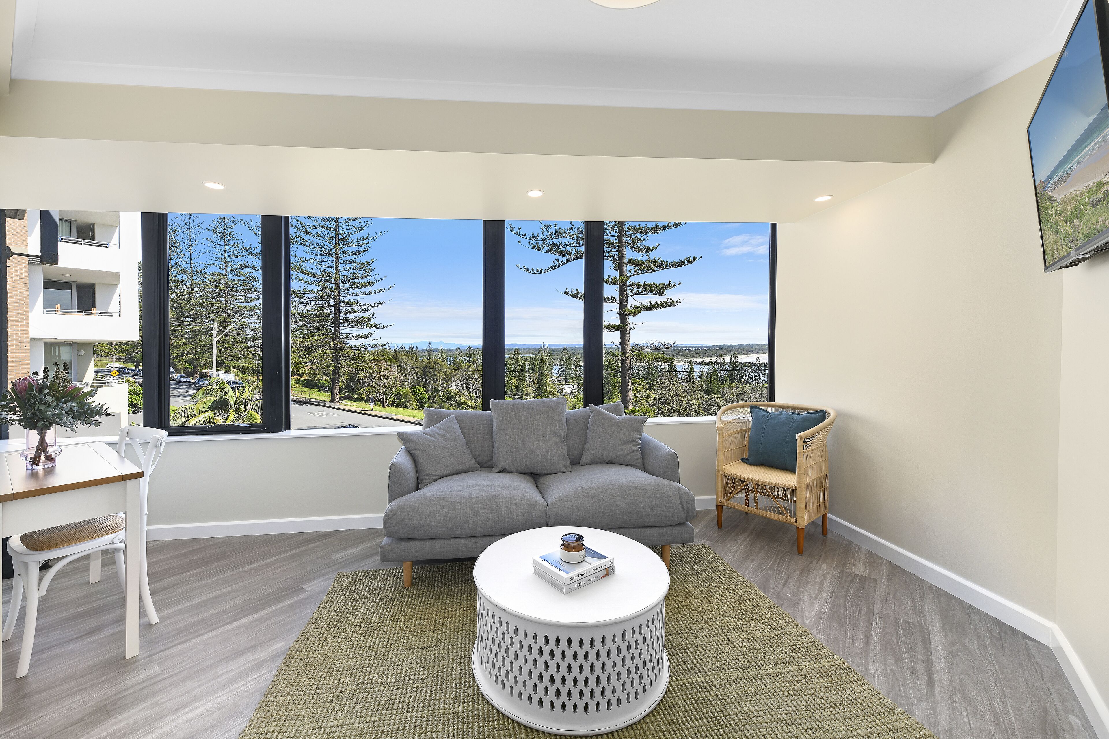 Uma sala de estar moderna com um sofá cinzento, uma mesa de centro redonda e um espaço de jantar com uma mesa e cadeiras brancas. Grandes janelas oferecem vista para árvores e uma paisagem distante.