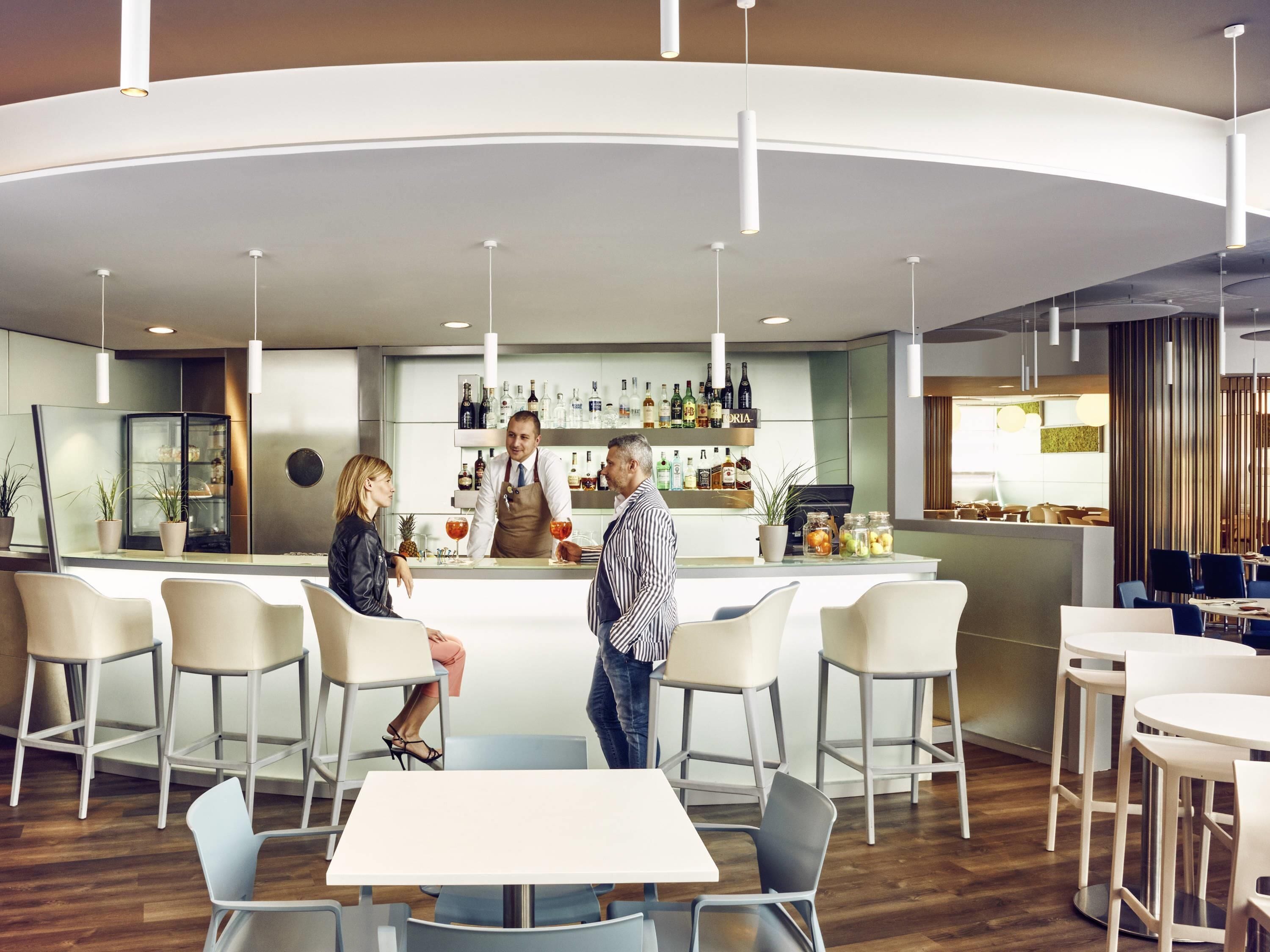 Uma área de bar moderna com teto curvo, banquetas e balcão com barman. Há mesas e cadeiras dispostas para refeições.