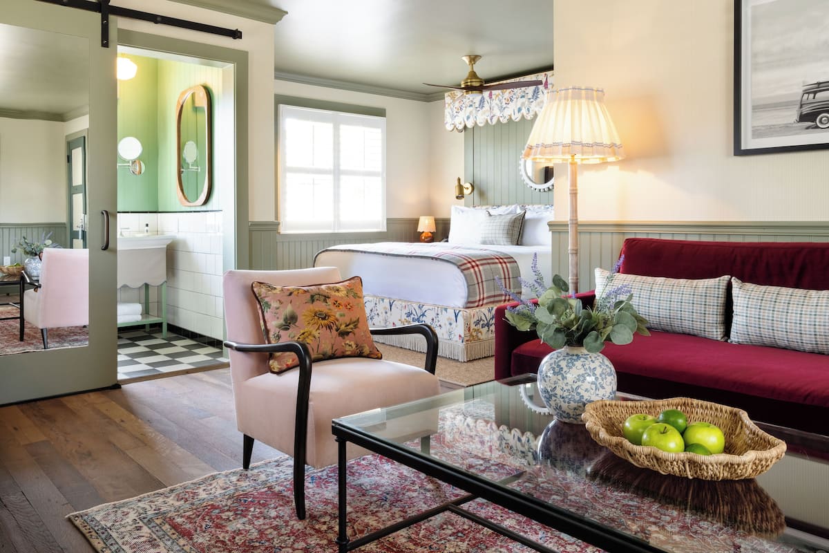 Um quarto aconchegante com uma cama, um sofá vermelho, uma mesa de centro de vidro com maçãs e uma cadeira com almofada floral.