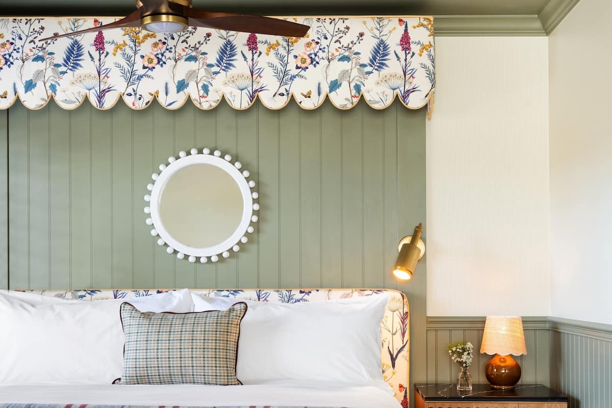Quarto com cama grande, espelho redondo, mesa de cabeceira com abajur e cabeceira com padrão floral.