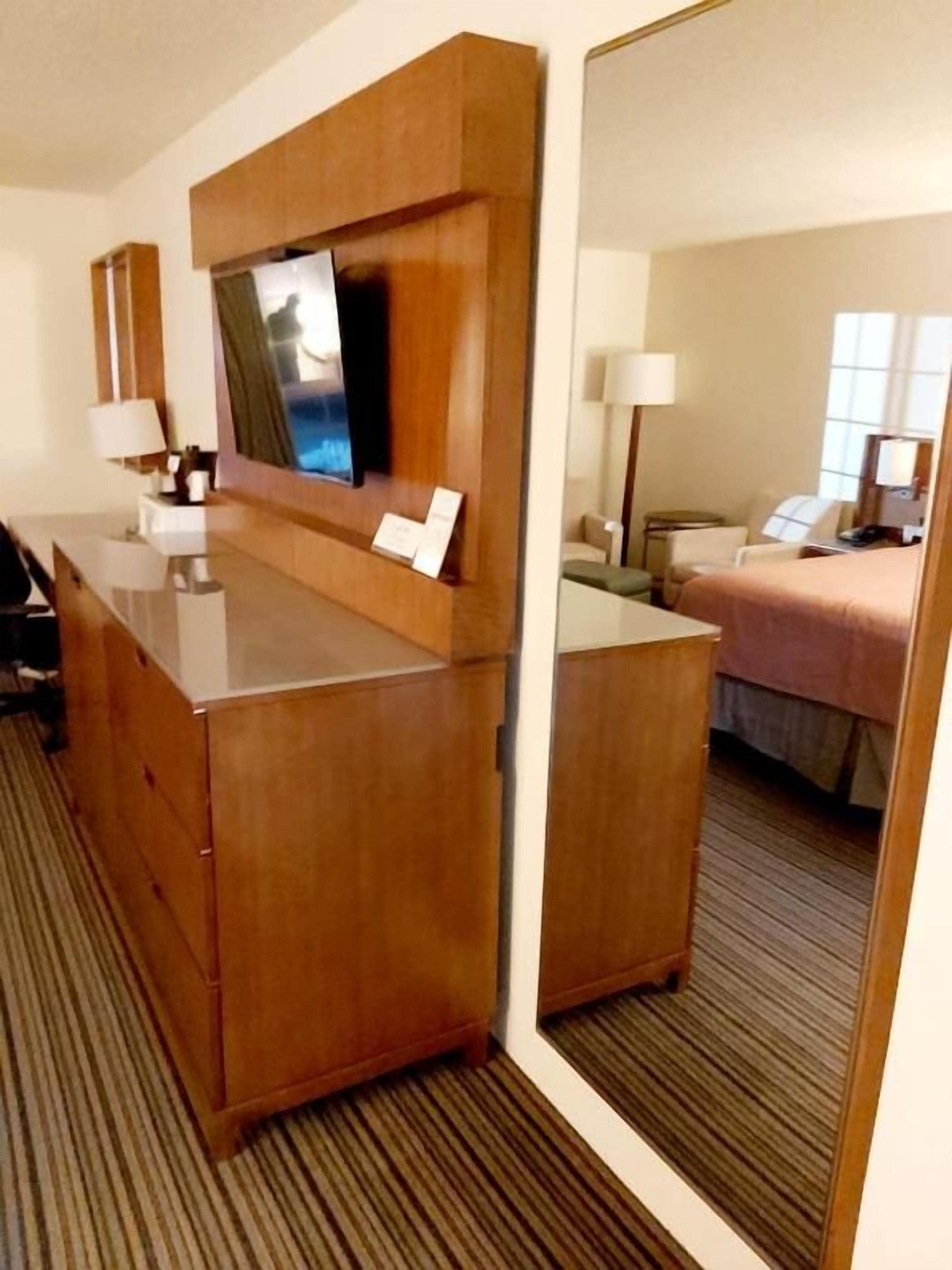 Quarto de hotel com penteadeira de madeira, televisão de tela plana, cama com manta rosa e espelho refletindo o quarto.