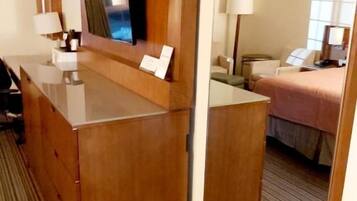 Quarto de hotel com penteadeira de madeira, televisão de tela plana, cama com manta rosa e espelho refletindo o quarto.