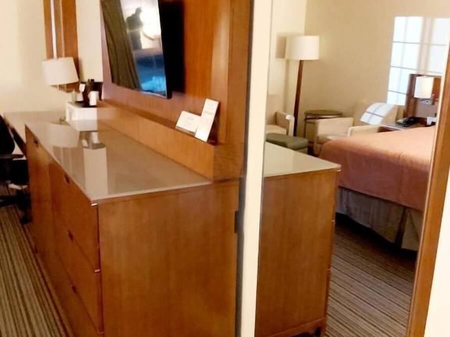 Quarto de hotel com penteadeira de madeira, televisão de tela plana, cama com manta rosa e espelho refletindo o quarto.