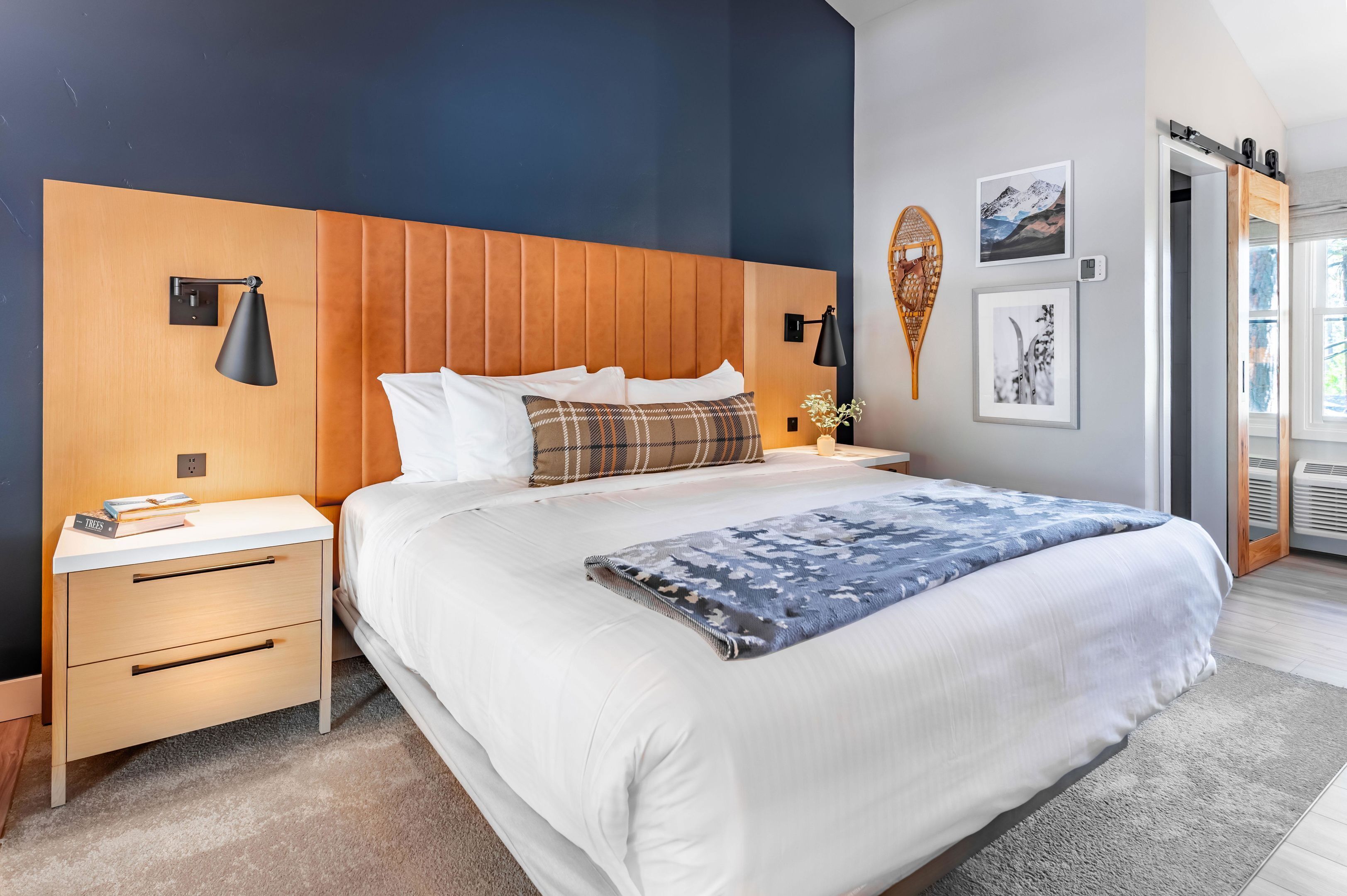 Um quarto moderno com cabeceira de madeira, uma cama grande com roupa de cama branca, uma mesinha de cabeceira com livros e luminárias fixadas na parede.