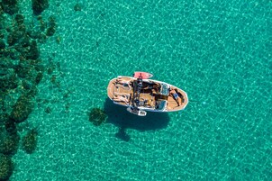 Vista aérea de um barco com cabine e área de convés, flutuando em águas turquesa claras com rochas visíveis submersas.