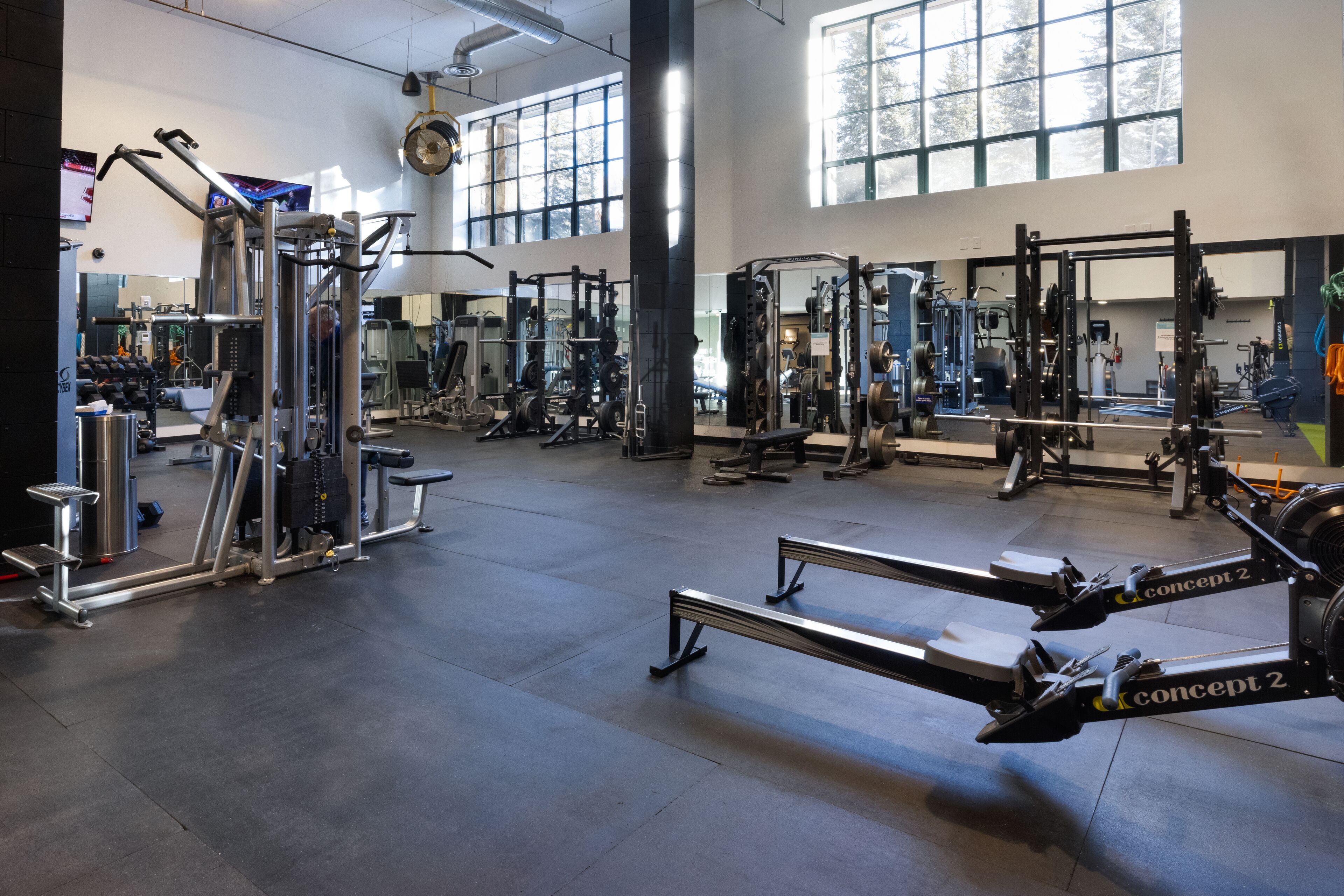Academia com diversos equipamentos de exercício, amplas janelas e pé direito alto.