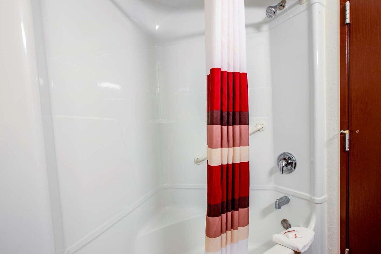 Um chuveiro no canto com parede revestida de azulejos brancos, cortina de chuveiro listrada em vermelho e bege, e uma pia pequena branca com um espelho.