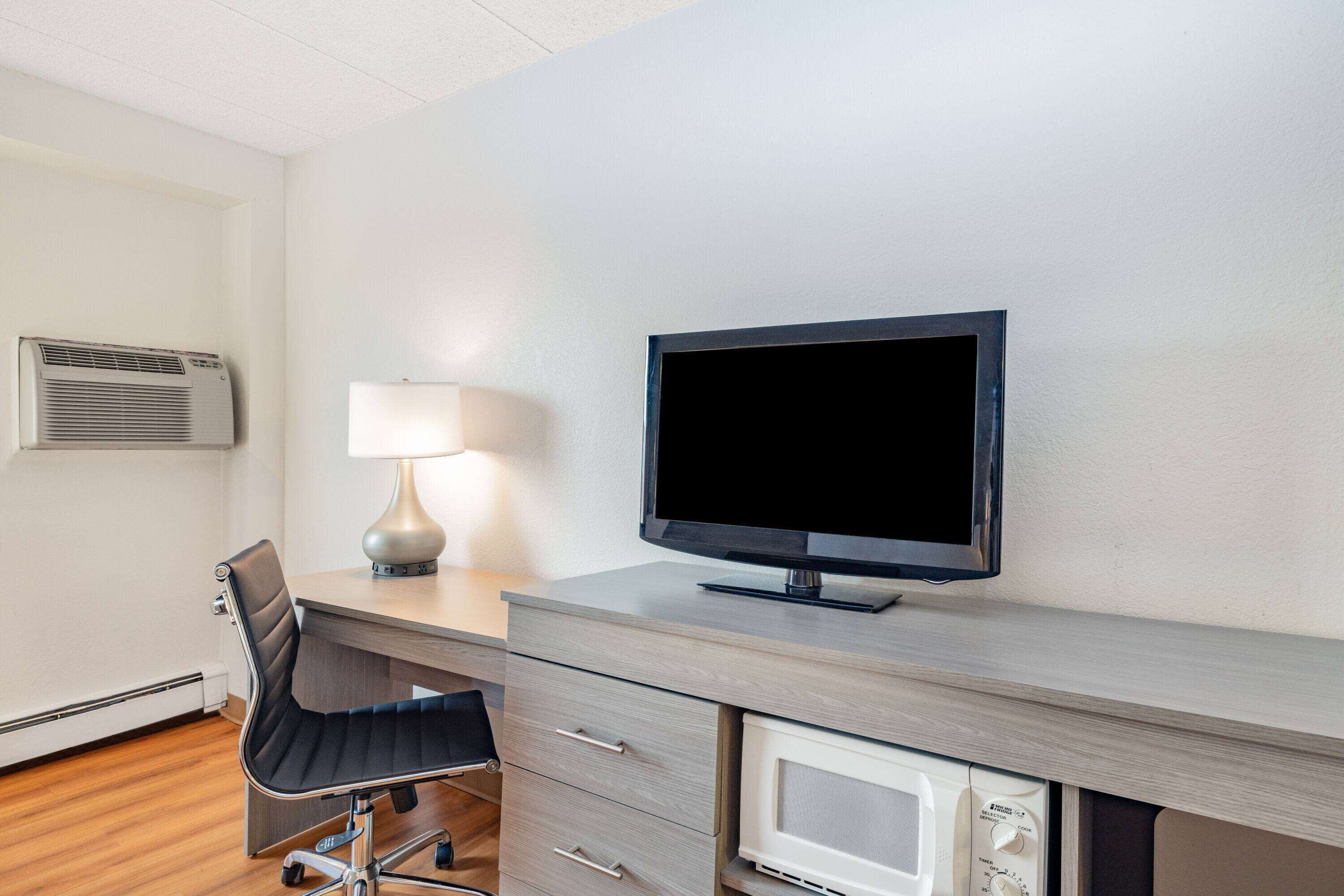 Quarto de hotel com escrivaninha, cadeira, televisão, luminária e micro-ondas.