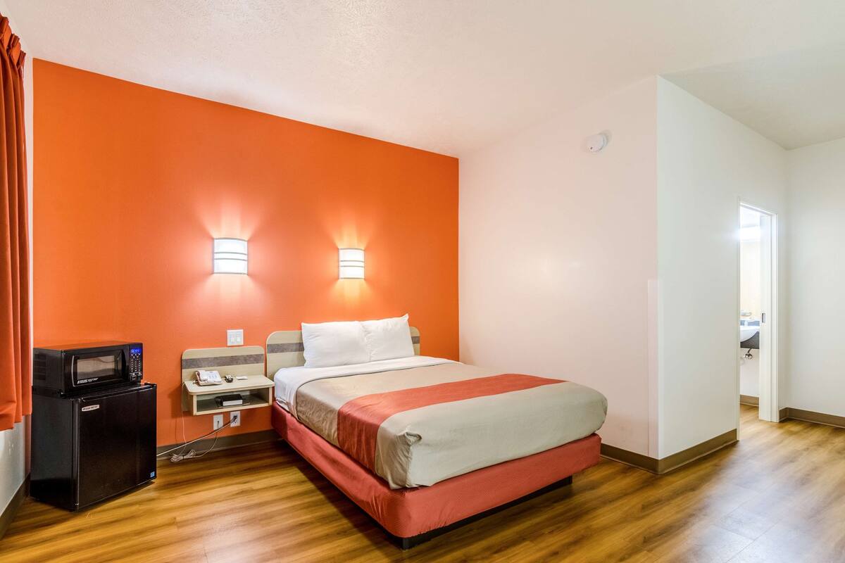 Quarto de hotel com uma cama, uma pequena escrivaninha com luminária, micro-ondas e cortina vermelha.