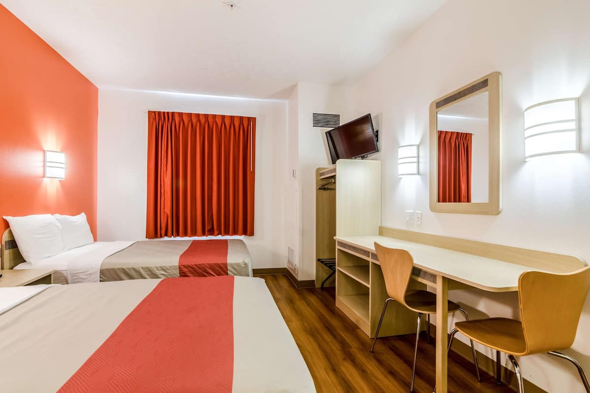 Quarto de hotel com duas camas, uma escrivaninha, uma cadeira, uma televisão e um espelho.