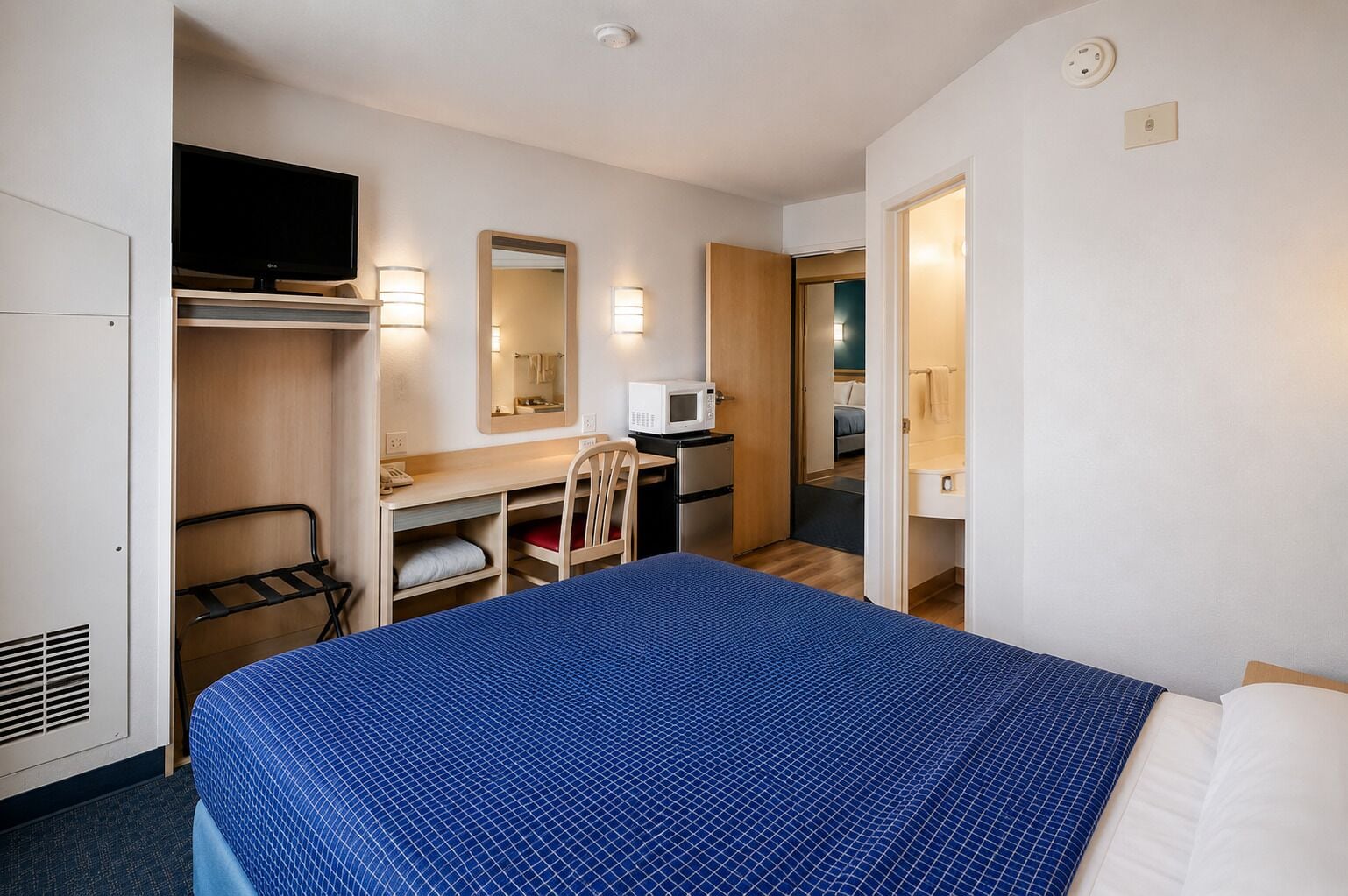 Quarto de hotel com cama, escrivaninha, poltrona, micro-ondas, televisão e guarda-roupa.