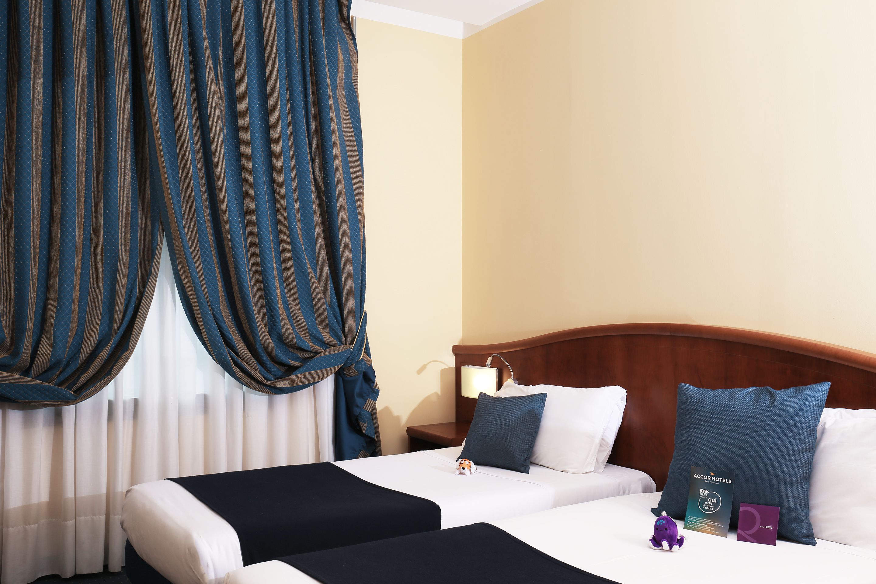 Quarto de hotel com duas camas, uma cabeceira de madeira, mesa de cabeceira com abajur e janela com cortinas.