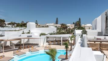 Uma área de piscina no terraço com espreguiçadeiras e uma mesa de jantar, cercada por edifícios brancos e palmeiras.