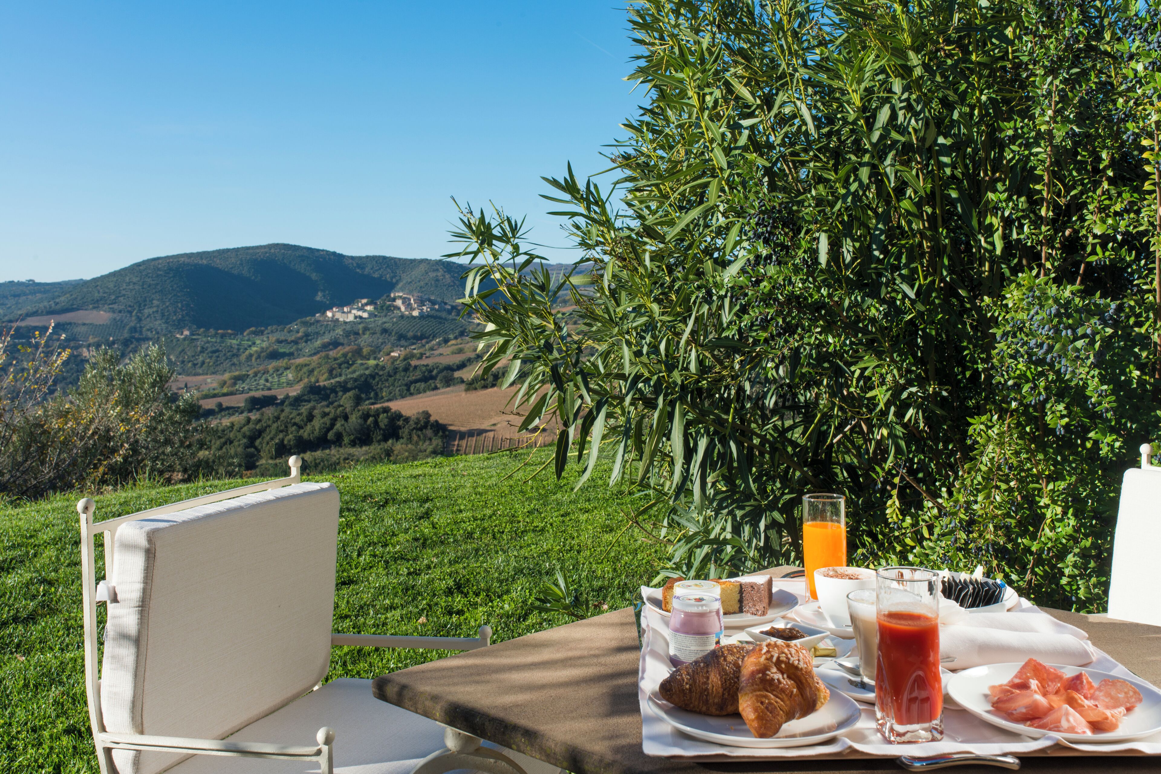 Mesa posta ao ar livre com vista para colinas ondulantes, uma cadeira e uma mesa de café da manhã com croissants, queijo e suco.