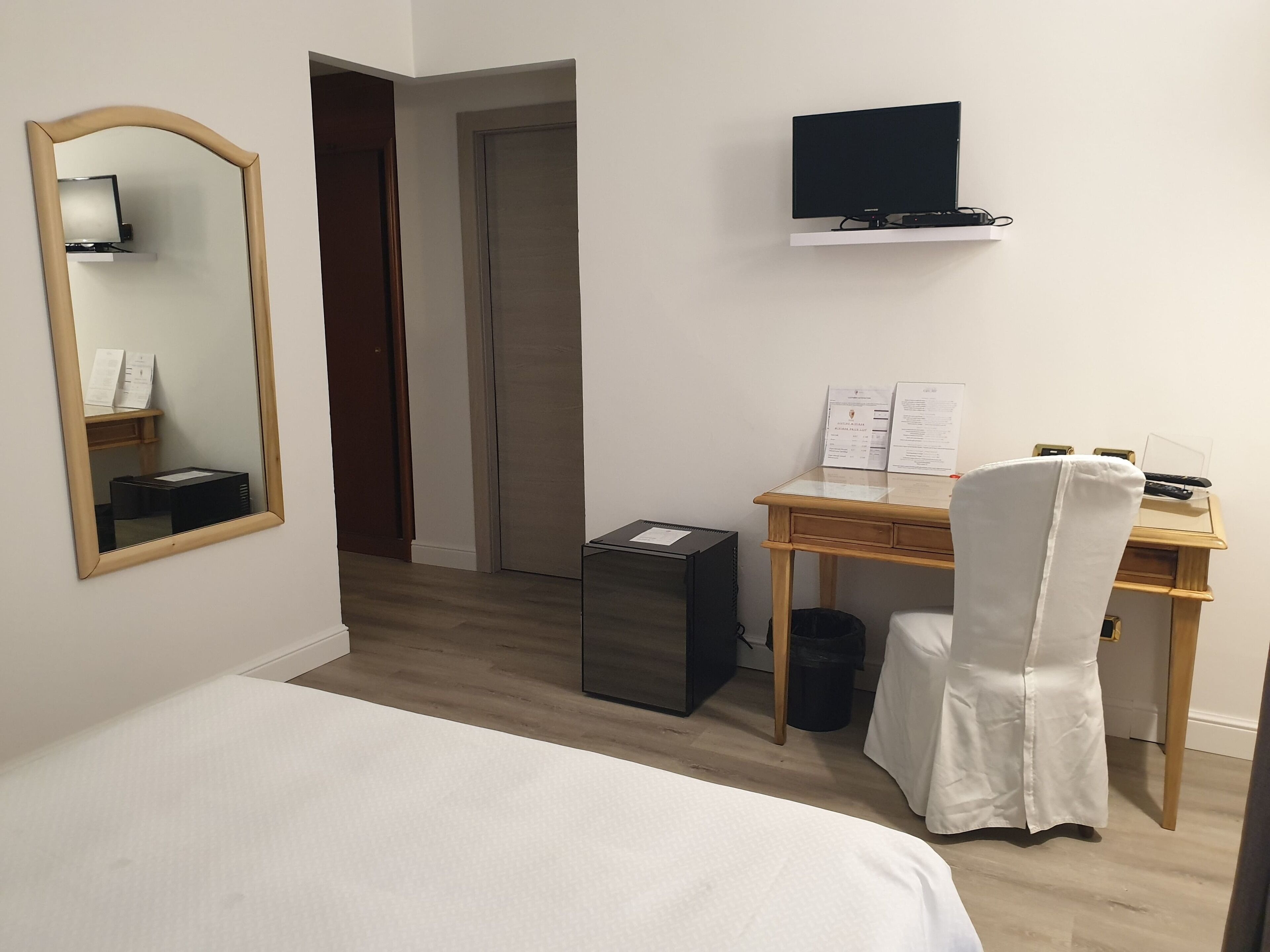 Quarto de hotel com cama, escrivaninha, cadeira, televisão e espelho.