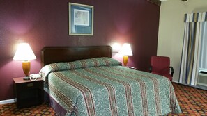 Quarto de hotel com uma cama grande, duas luminárias laterais, uma cadeira vermelha, um quadro na parede e um tapete estampado.