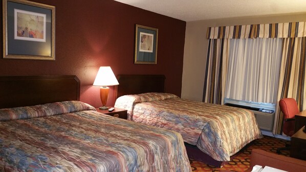 Quarto de hotel com duas camas, uma mesa de cabeceira com abajur, uma janela com cortinas listradas e um ar-condicionado fixado na parede.