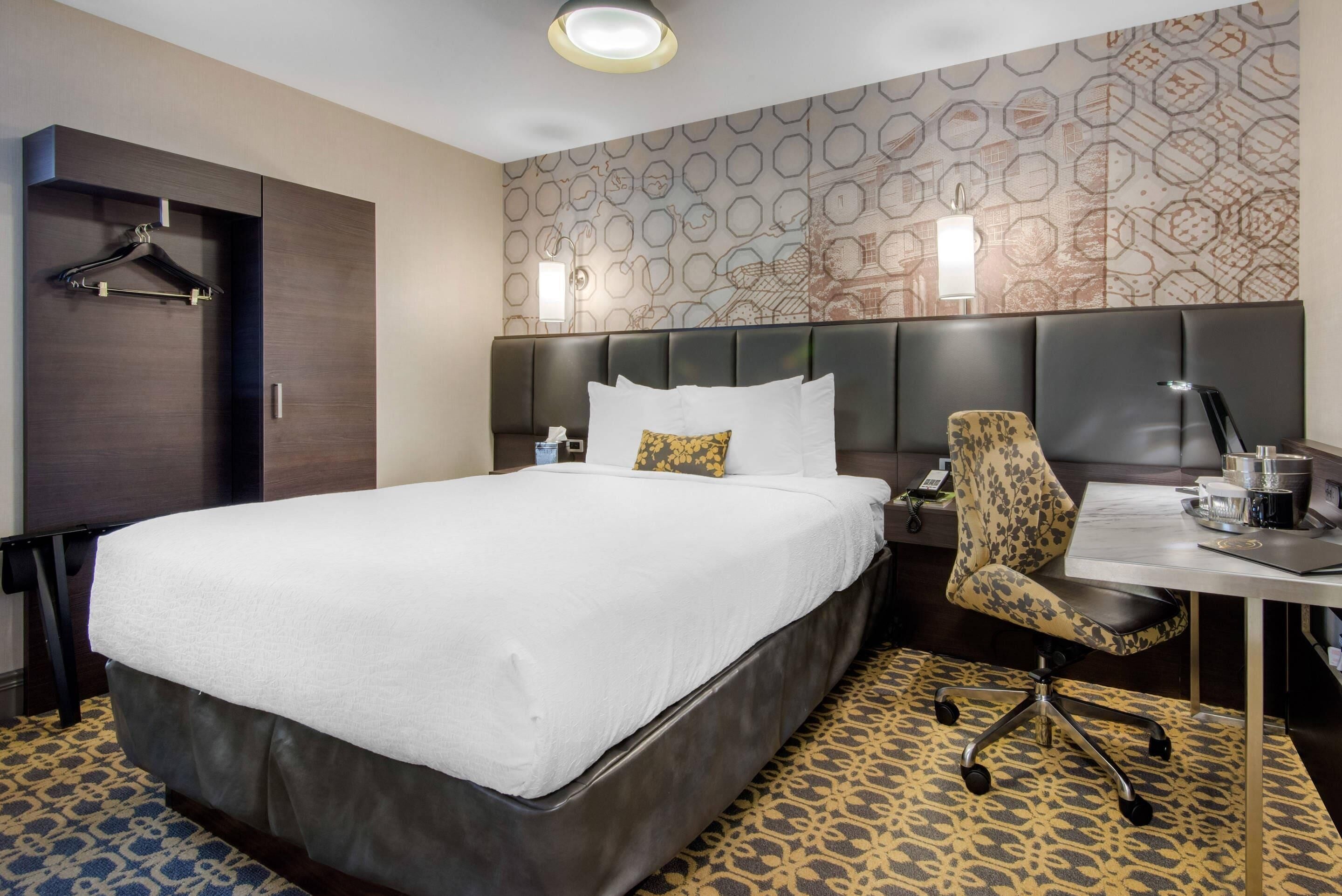 Quarto de hotel com uma cama grande, uma escrivaninha com cadeira, um guarda-roupa e uma parede estampada.