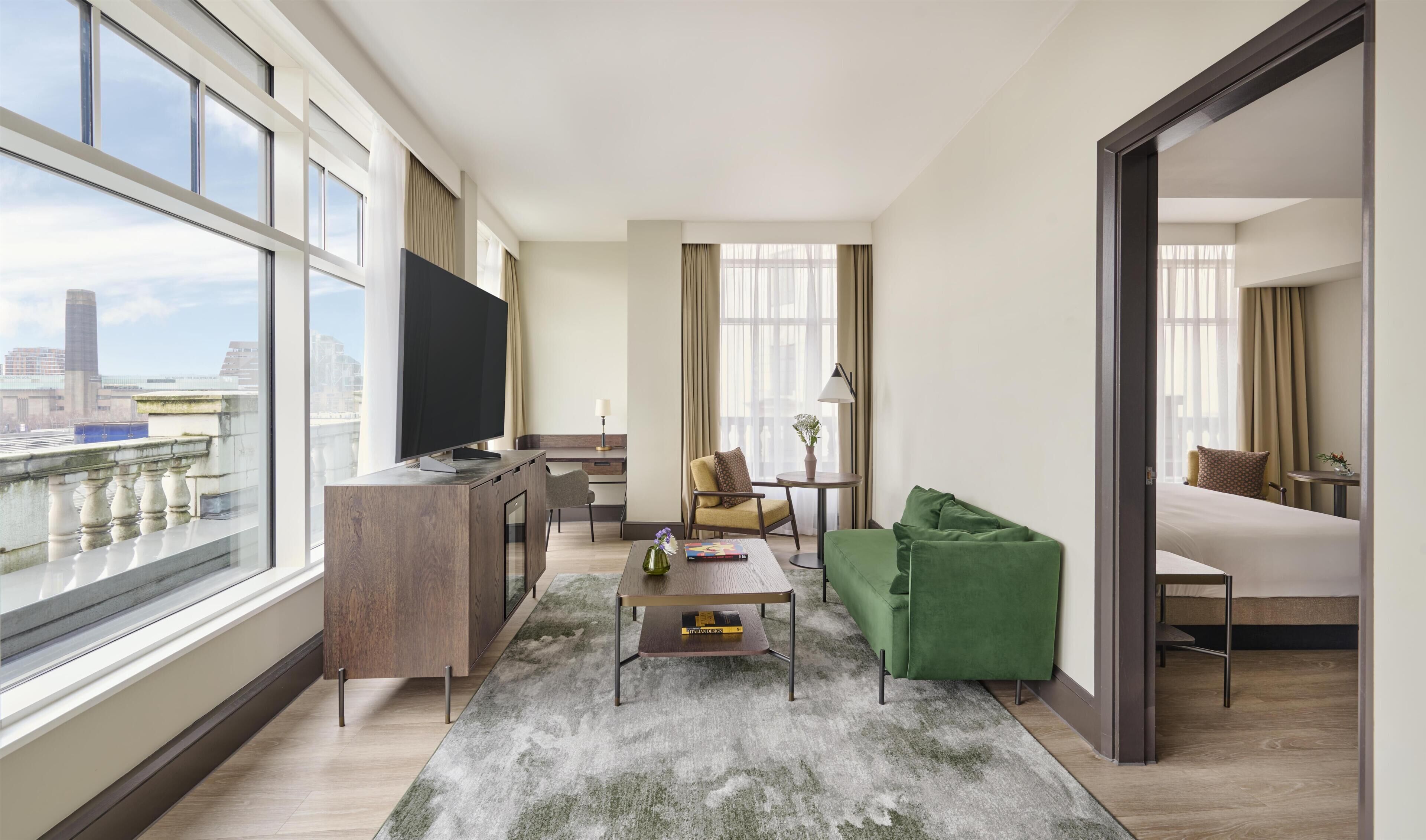 Quarto de hotel moderno com uma janela grande, televisão de tela plana, sofá verde, mesa pequena e uma cama.