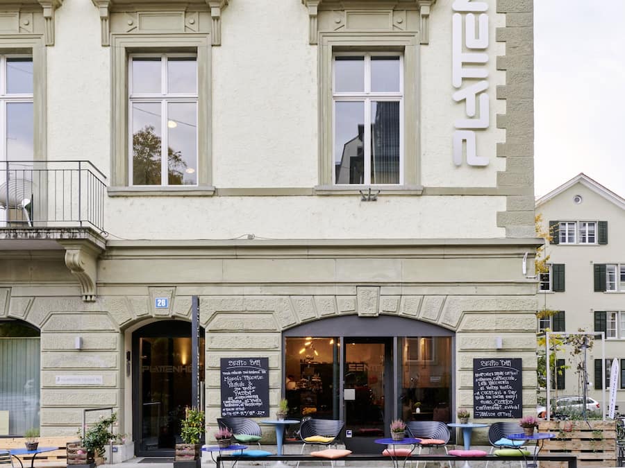 Um edifício com fachada clássica, um café no andar térreo com mesas ao ar livre e uma entrada visível.