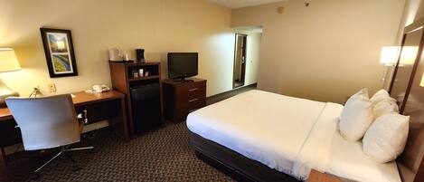 Quarto de hotel com cama, escrivaninha, cadeira, televisão e um pequeno frigorífico.