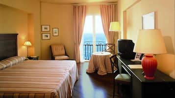 Quarto de hotel com uma cama, uma secretária com televisão, uma cadeira, uma mesa pequena e uma varanda com cortinas.