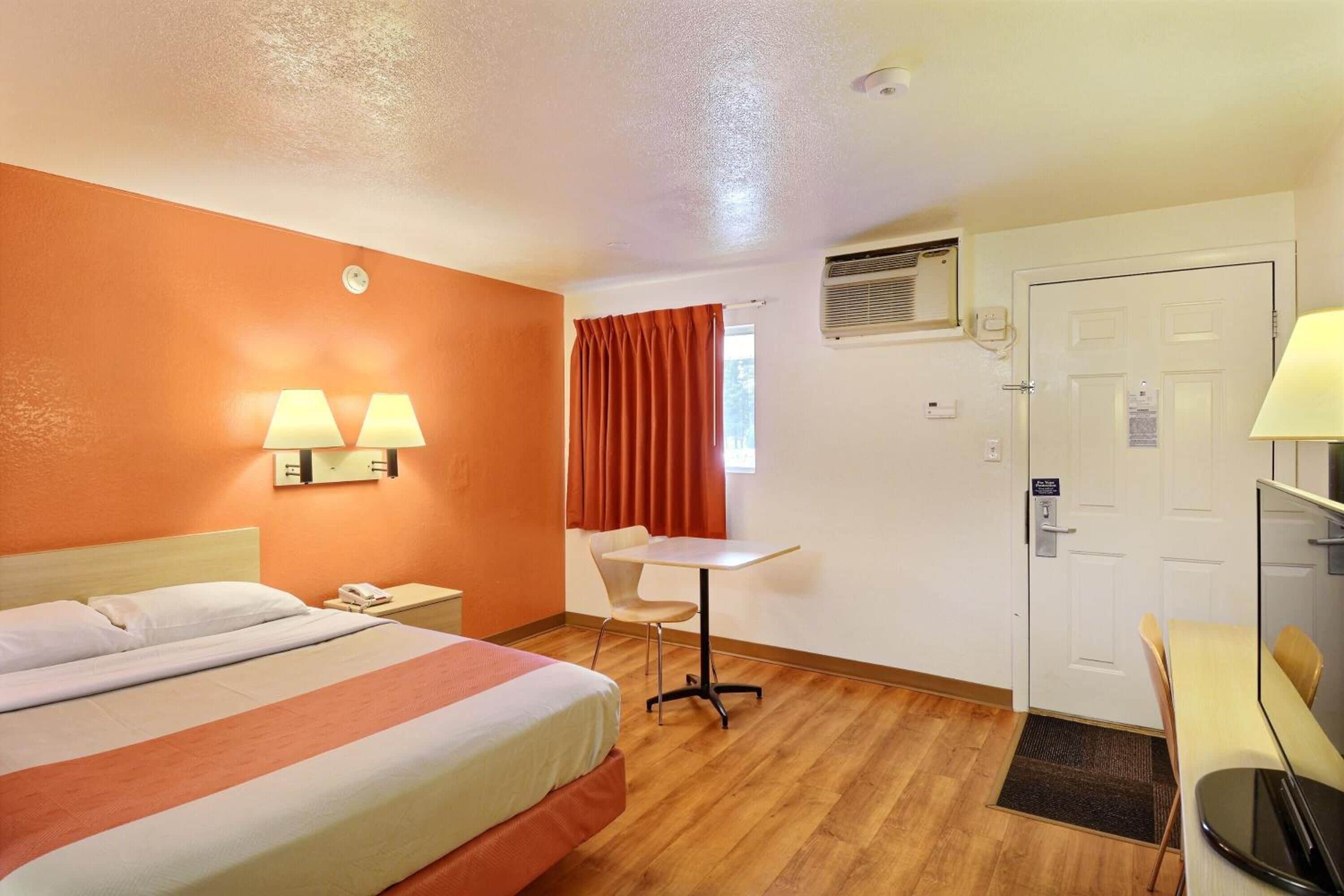 Quarto de hotel com cama, duas luminárias de cabeceira, escrivaninha com cadeira, porta e ar condicionado.
